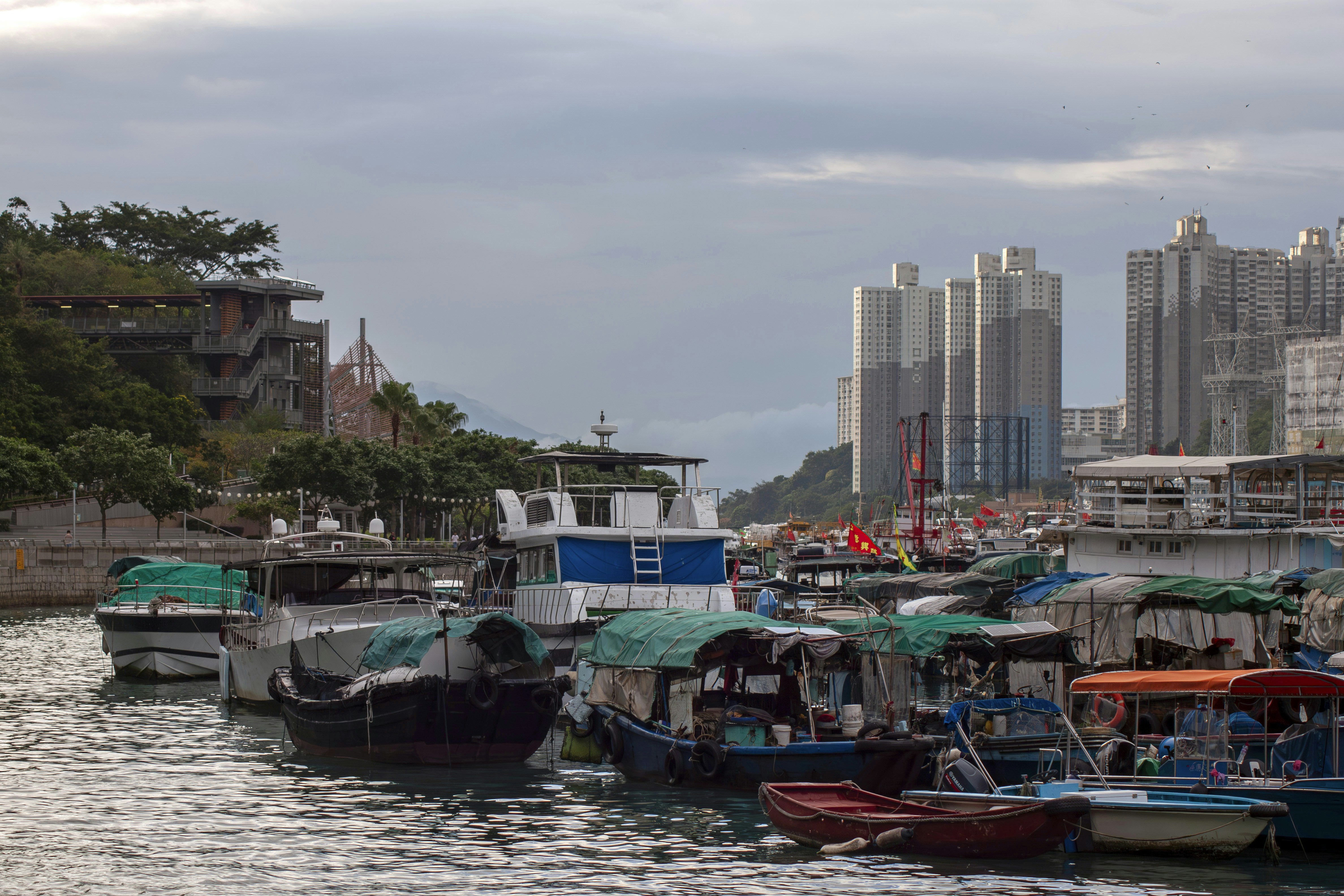 Boats docked along a bustling harbor with tall city buildings in the background under a cloudy sky.