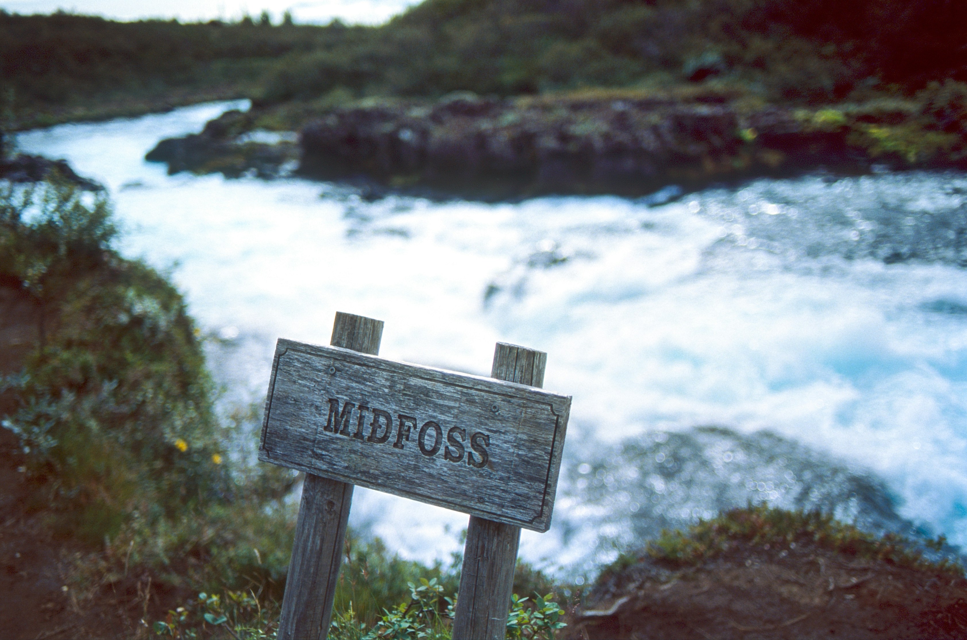 Weathered wooden sign reading MIDFOSS leans toward a foaming river, framed by mossy banks and rocky scrub.
