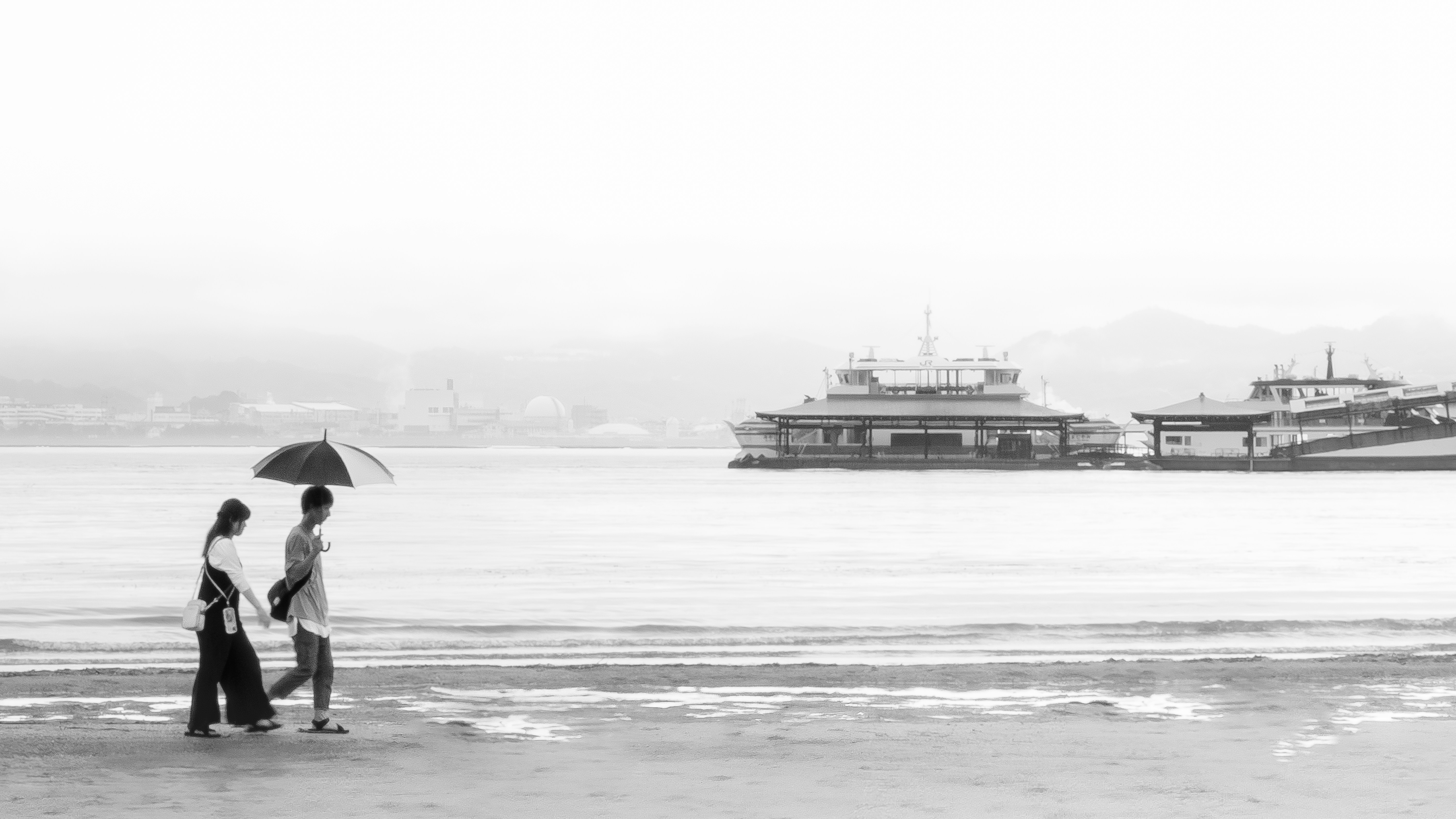 Two individuals walking under an umbrella along a misty shoreline with ferries in the distance.