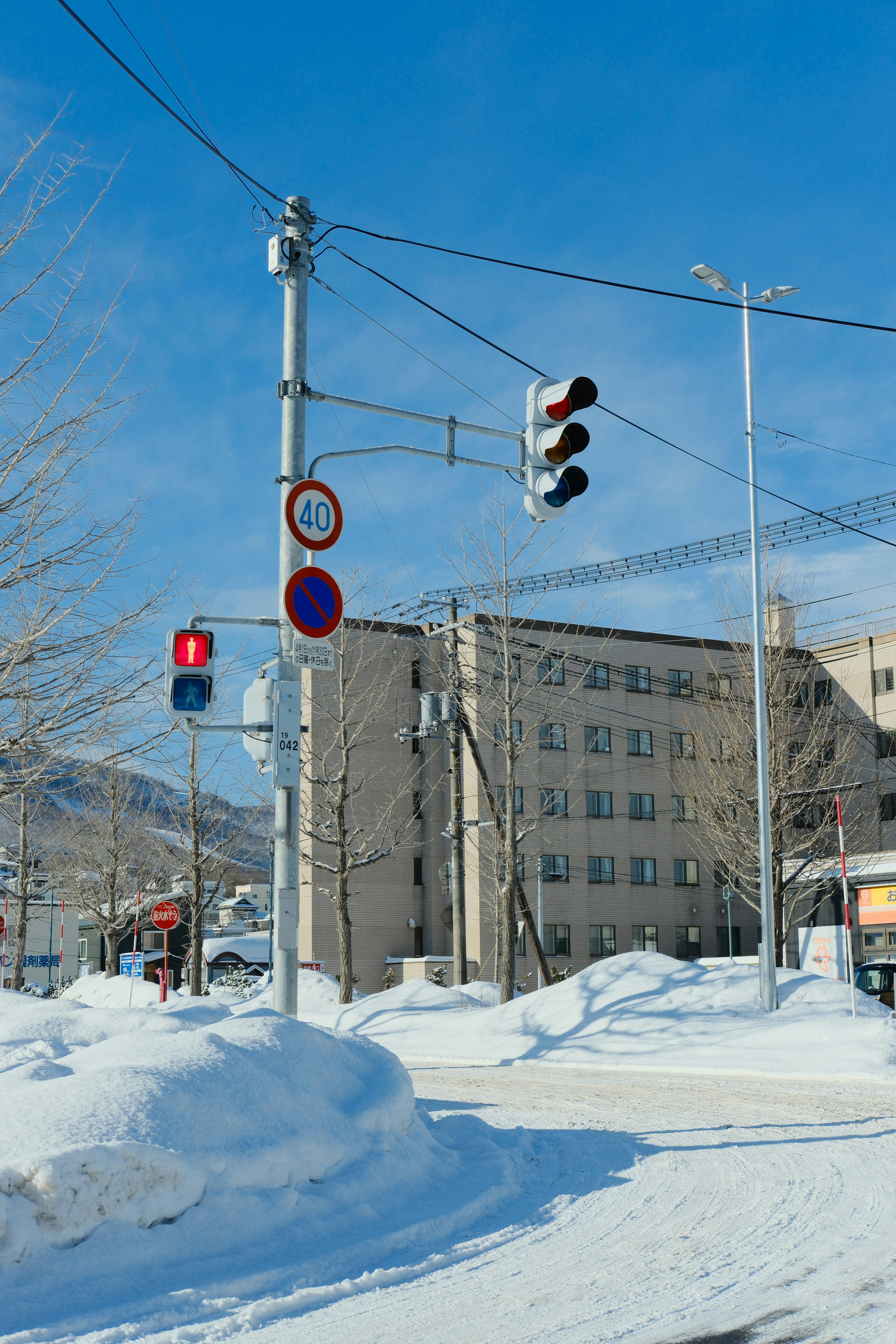 Photograph of a winter street scene with snowbanks along the curb, traffic lights, and road signs set against a crisp blue sky. A gray building anchors the background.