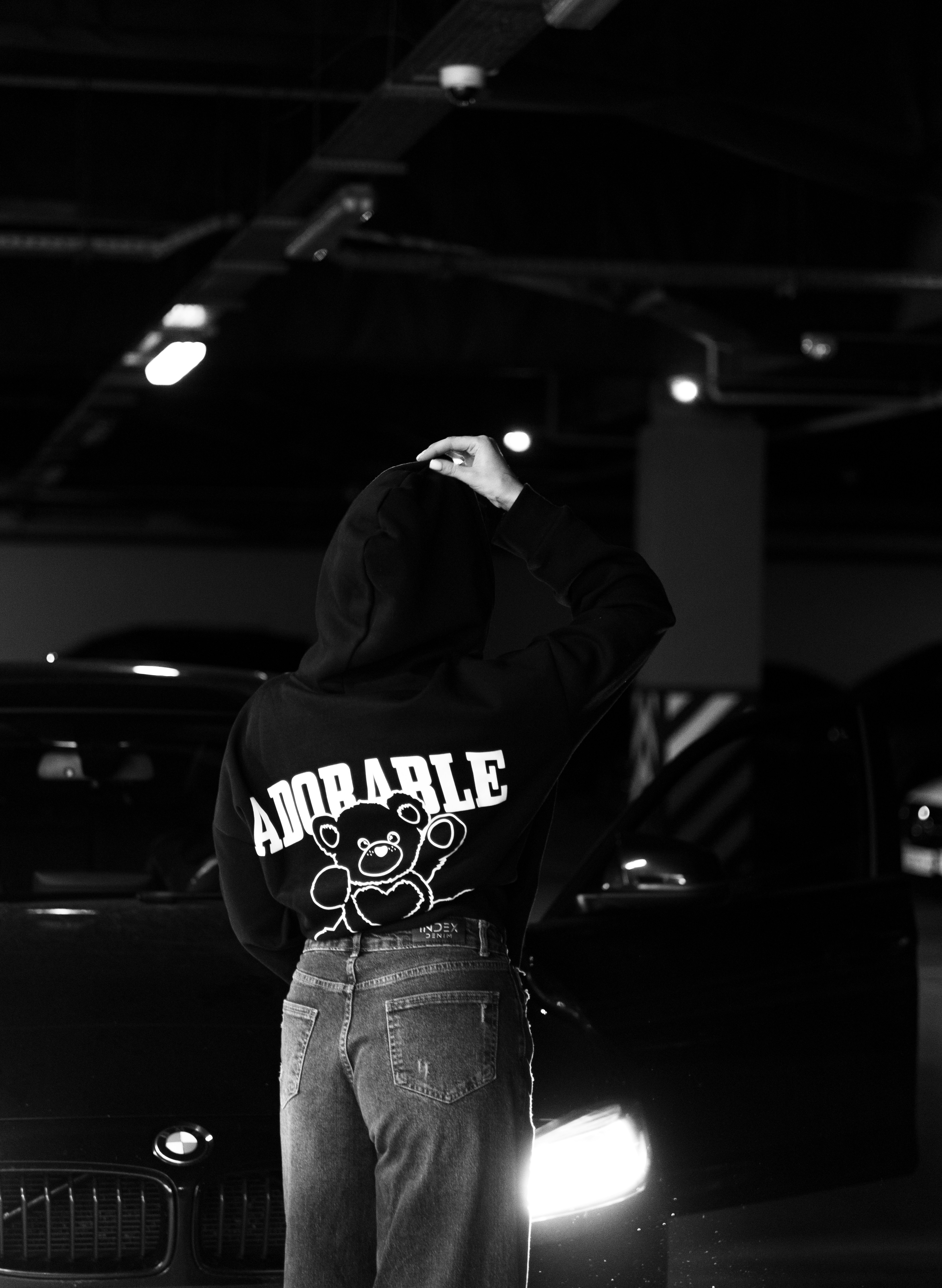 Person in a black hoodie with 'ADORABLE' text and bear illustration, standing beside a car in a dimly lit parking structure.