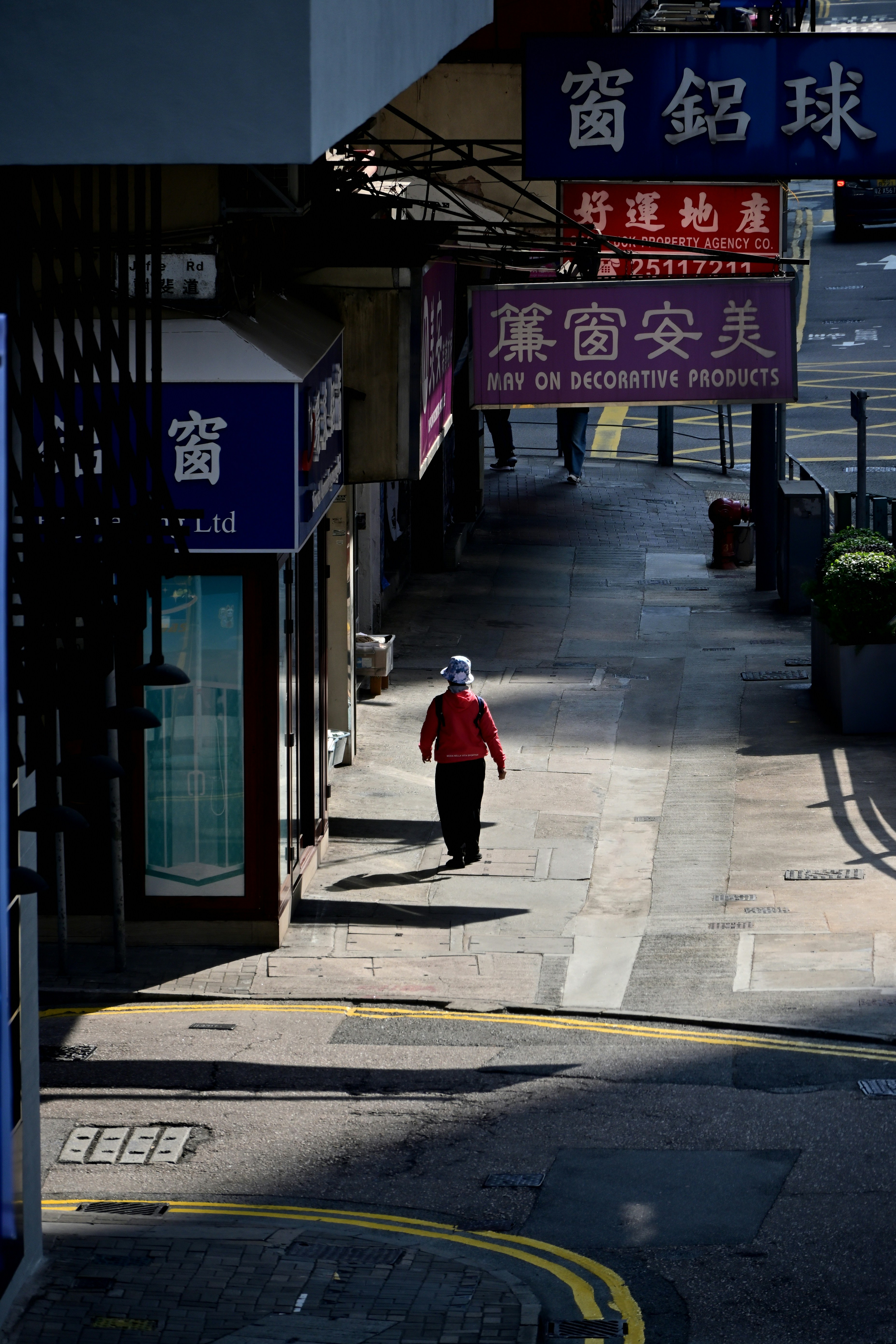 Person in red shirt walking through shadowed city street with colorful signage overhead.