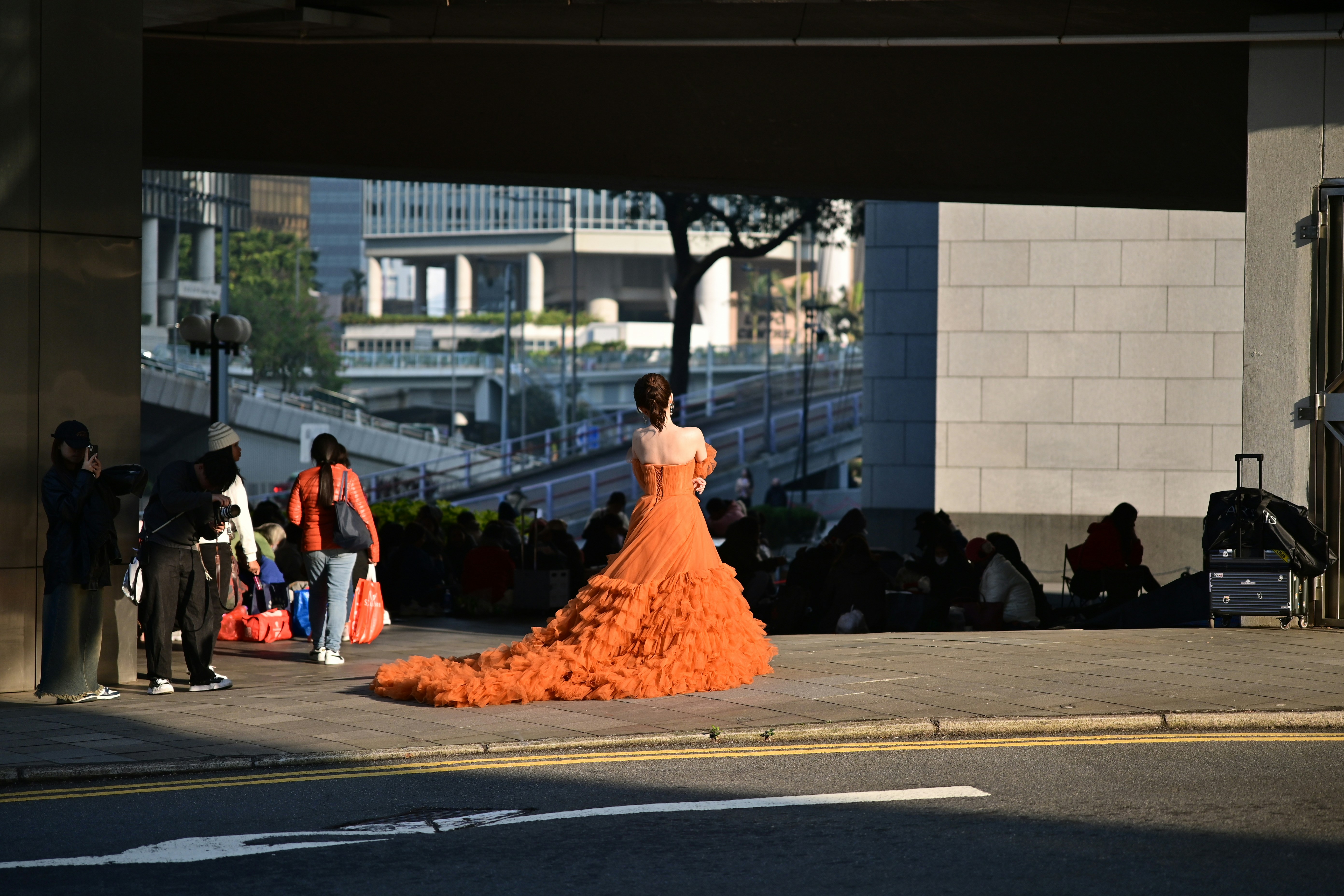 Woman in an elaborate orange dress walks under a city overpass, contrasting with the urban backdrop.