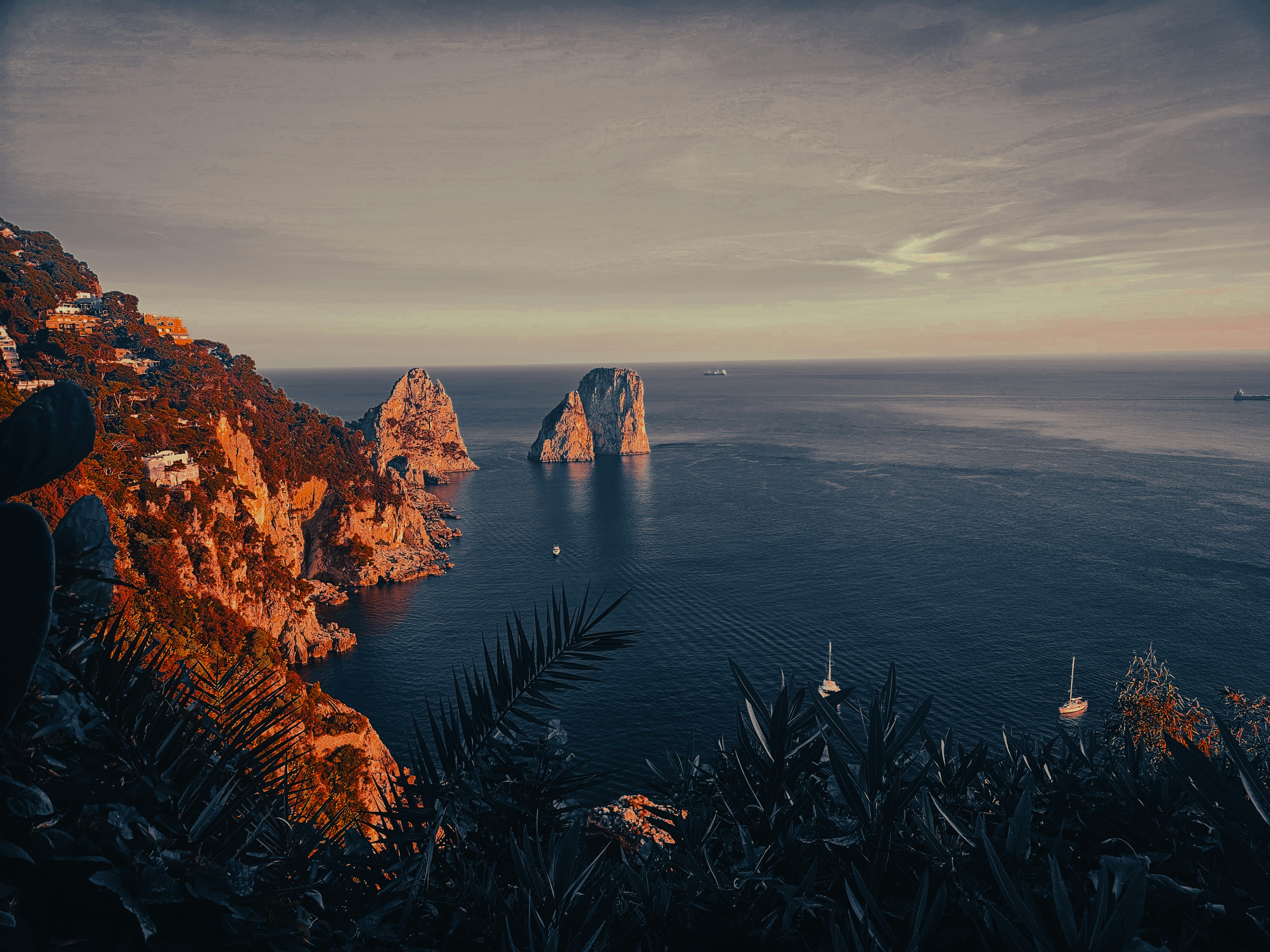 Coastal landscape photograph featuring orange cliff faces, sea stacks rising from calm blue water, and a muted dusk sky.