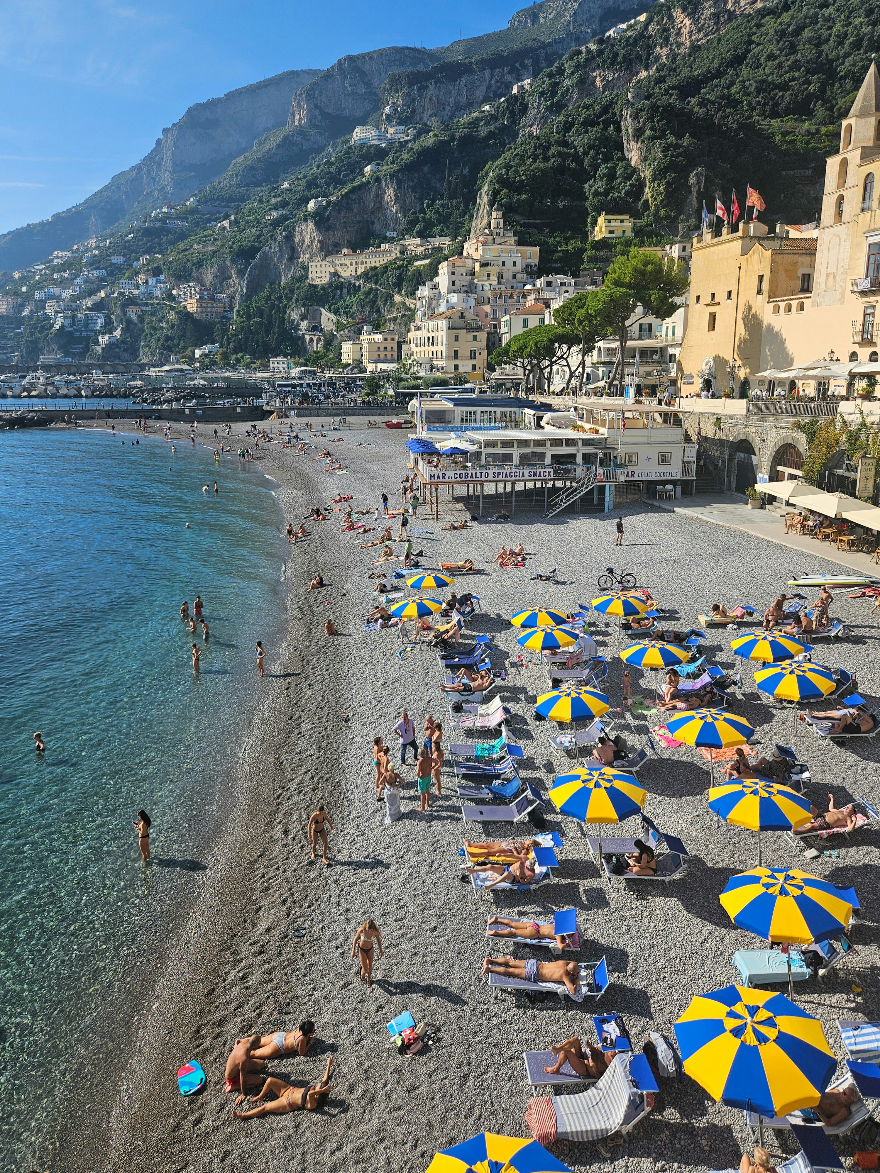 Vibrant beach scene with colorful umbrellas and sunbathers along a pebbled shore, backed by rugged coastal cliffs.