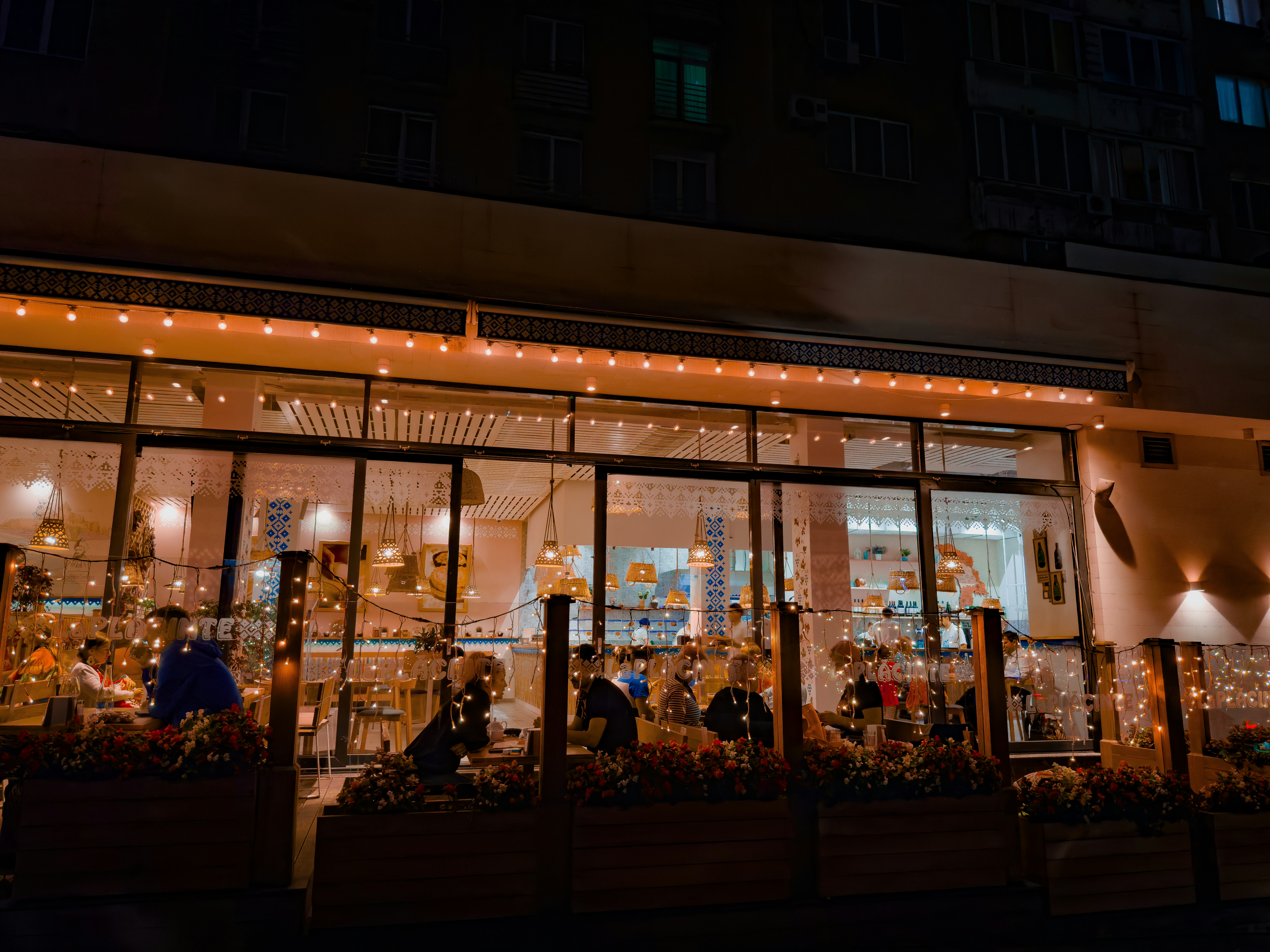 Warm lights illuminate a café's glass facade, showcasing an inviting interior with patrons inside.