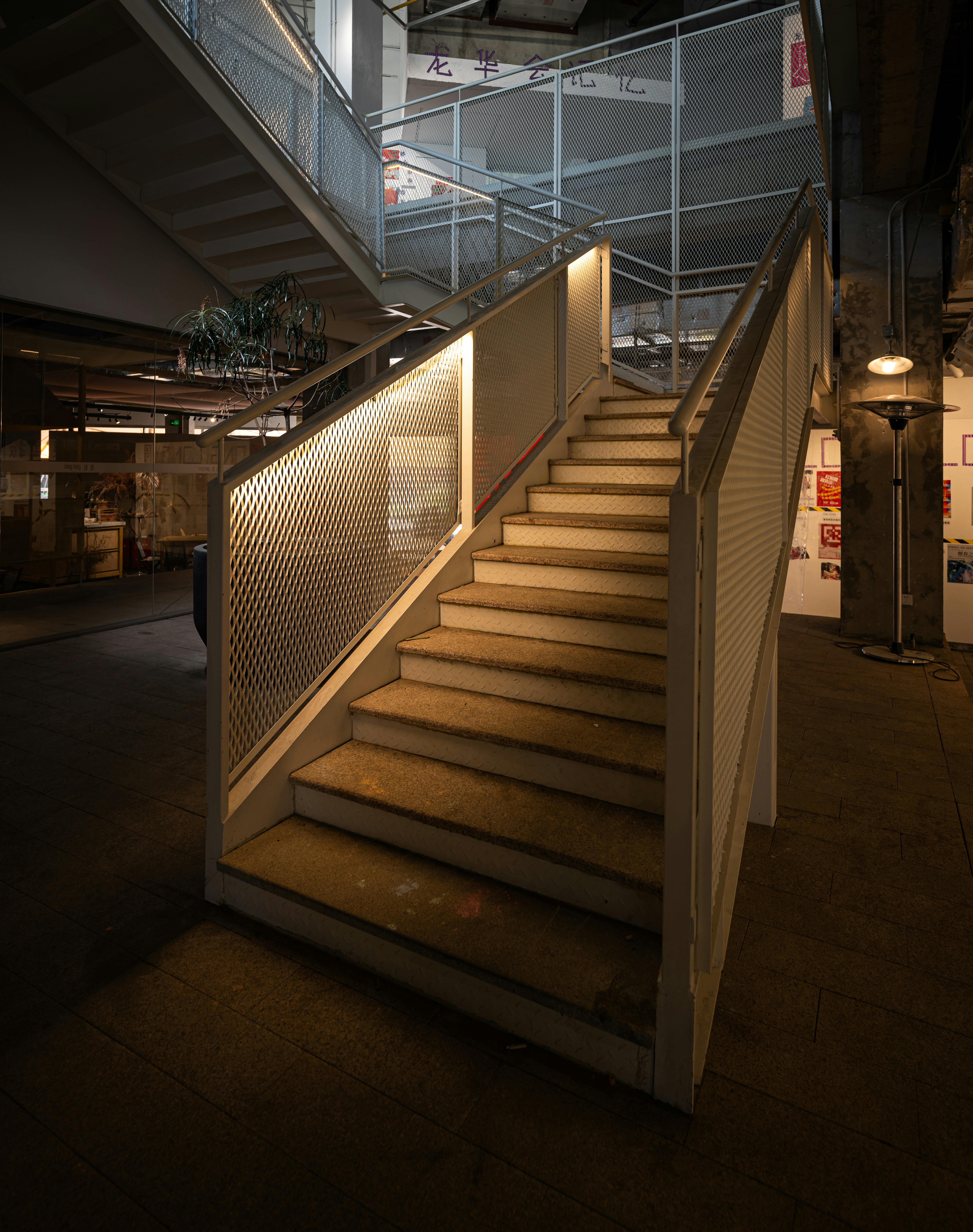 Metal stairway illuminated by warm ambient light in an industrial environment. Geometric patterns of mesh railing stand out against dim surroundings.