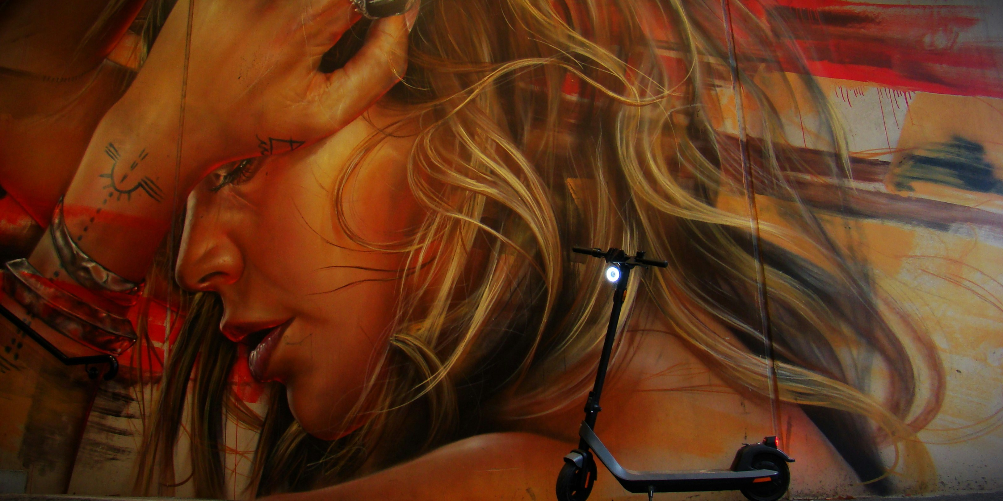 Vivid graffiti painting close-up of a woman's face with windswept blonde hair and bold red lips painted on a weathered wall.