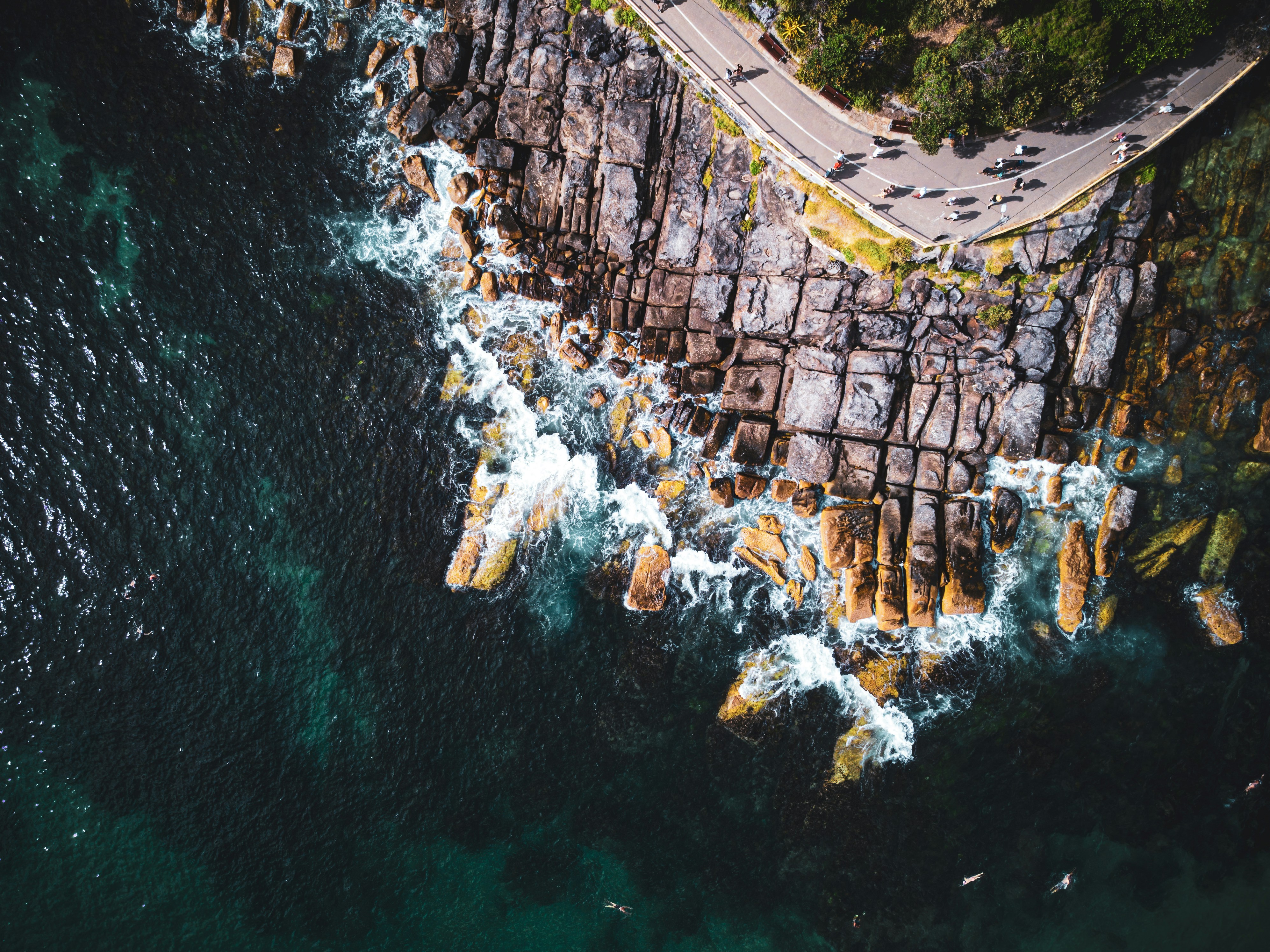 Aerial view of rugged cliffs meeting vibrant ocean waves with a sunlit pathway above.