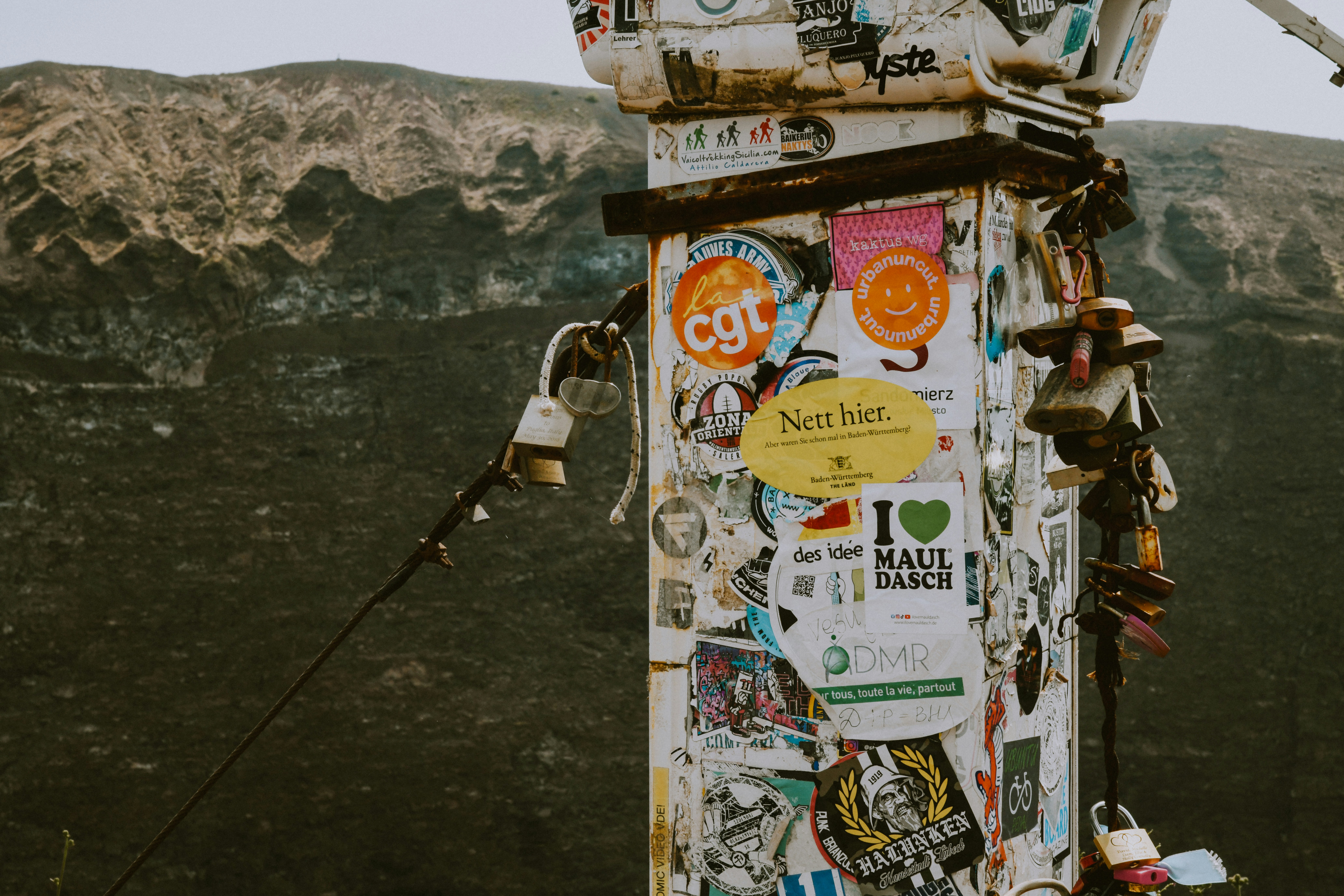 Wooden post adorned with colorful stickers and locks, set against a rugged mountainous backdrop.