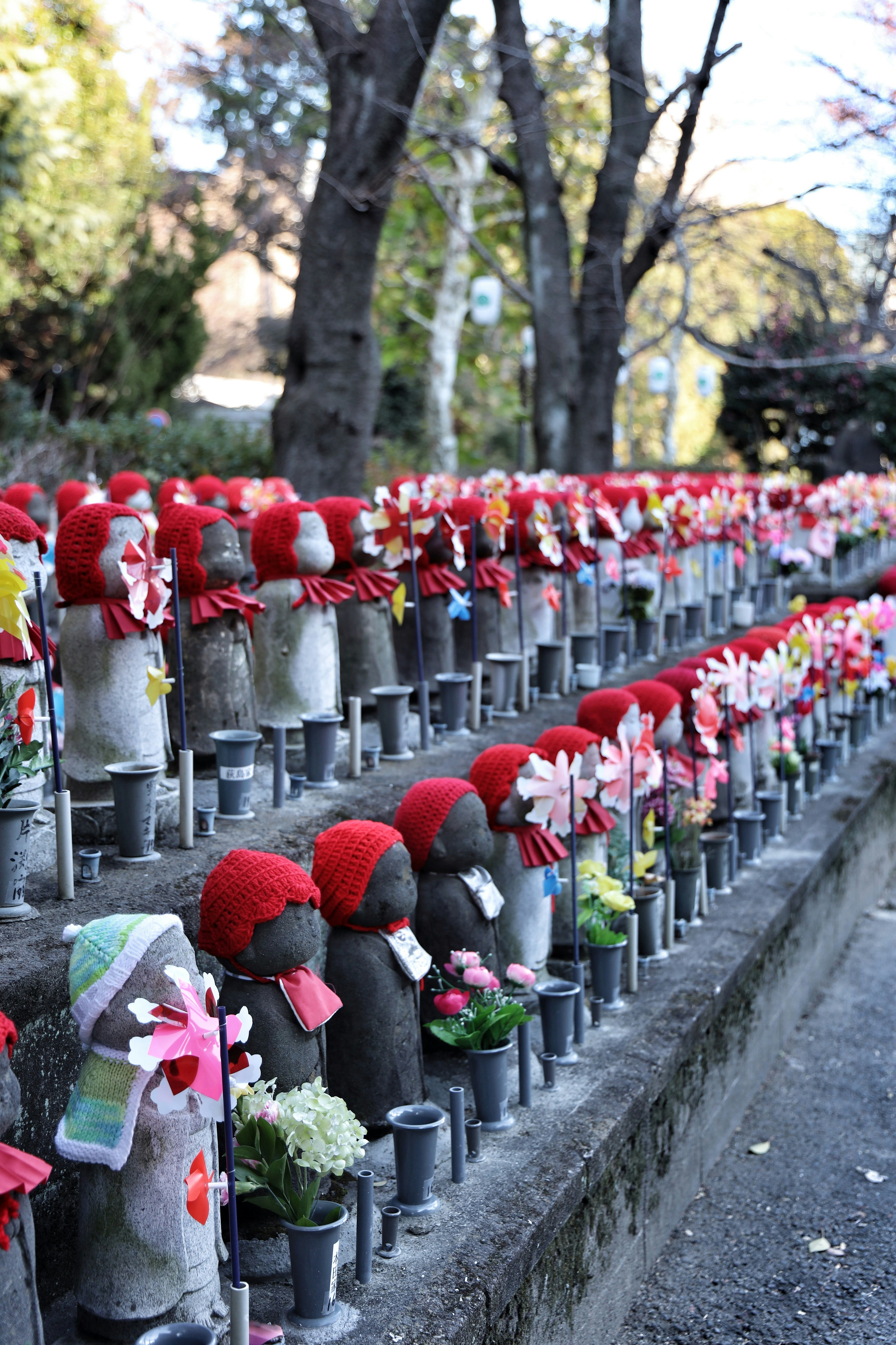 Rows of stone statues adorned with red hats and colorful flowers, symbolizing remembrance and care in a serene outdoor setting.