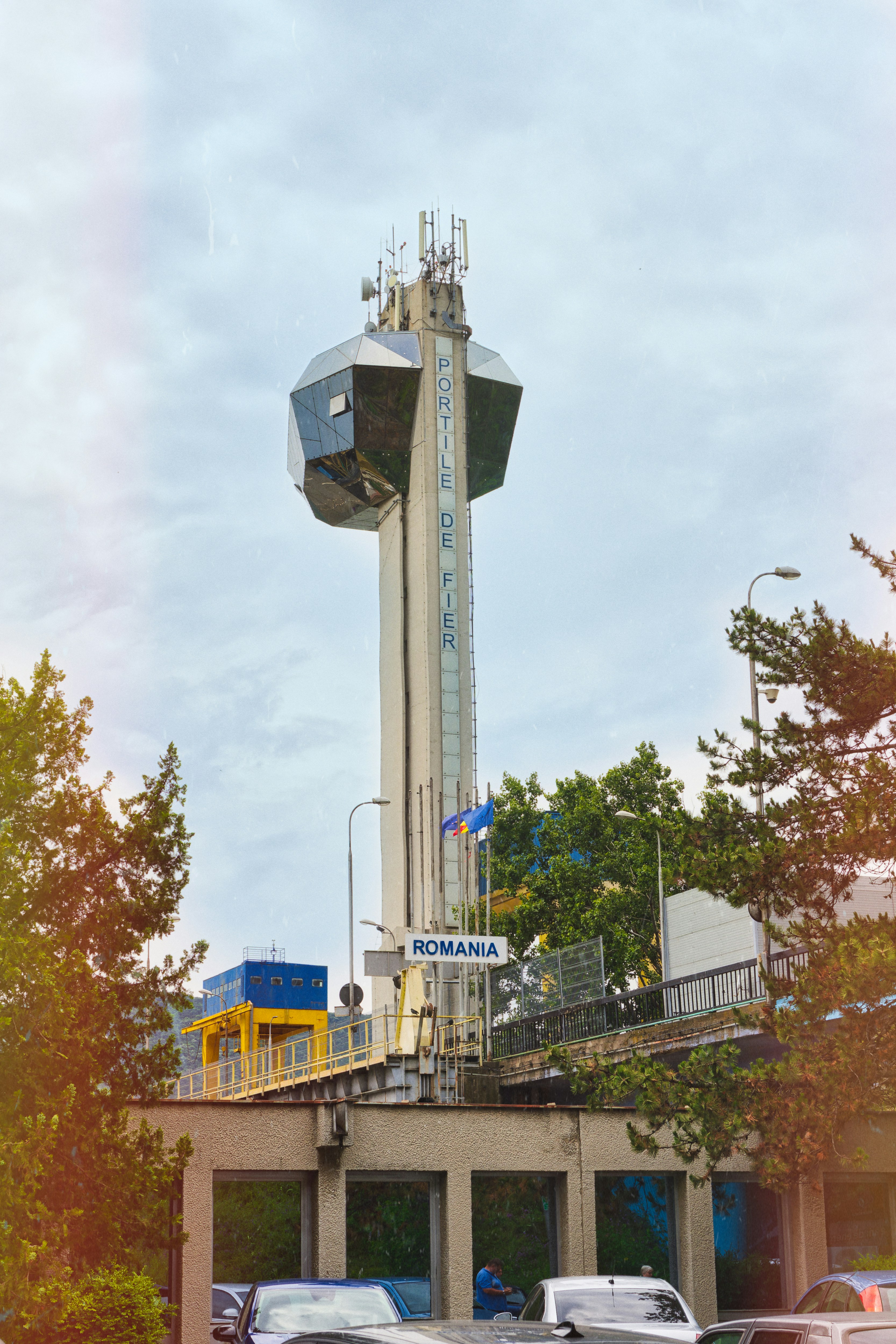 Tall observation tower with geometric design rises among trees and buildings under a cloudy sky.