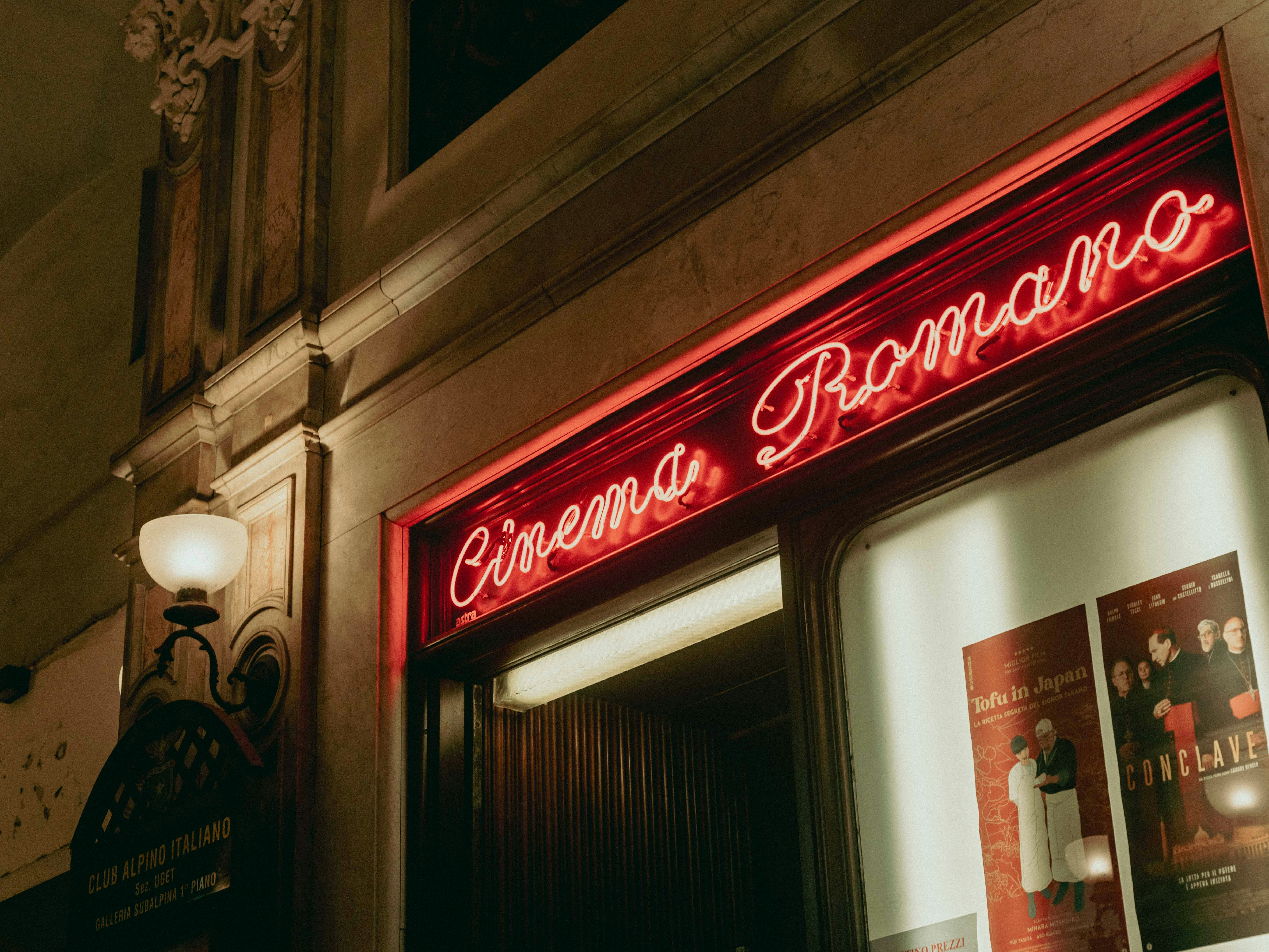 Illuminated vintage sign of Cinema Romano, showcasing a warm glow against the evening backdrop. Movie posters add to the nostalgic ambiance.
