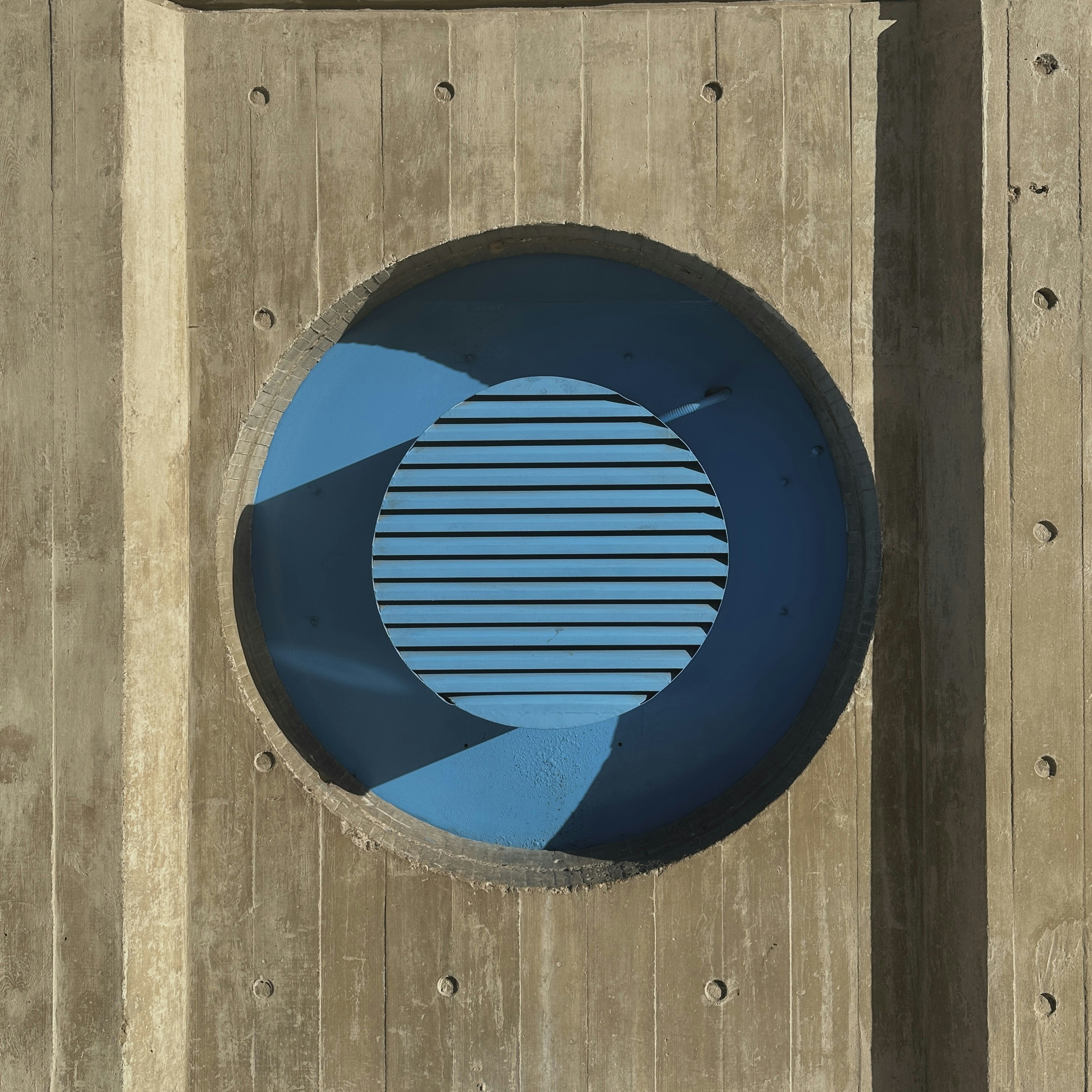 Blue Circular Vent in a Brutalist Concrete Frame: Geometry of Slats and Shadow