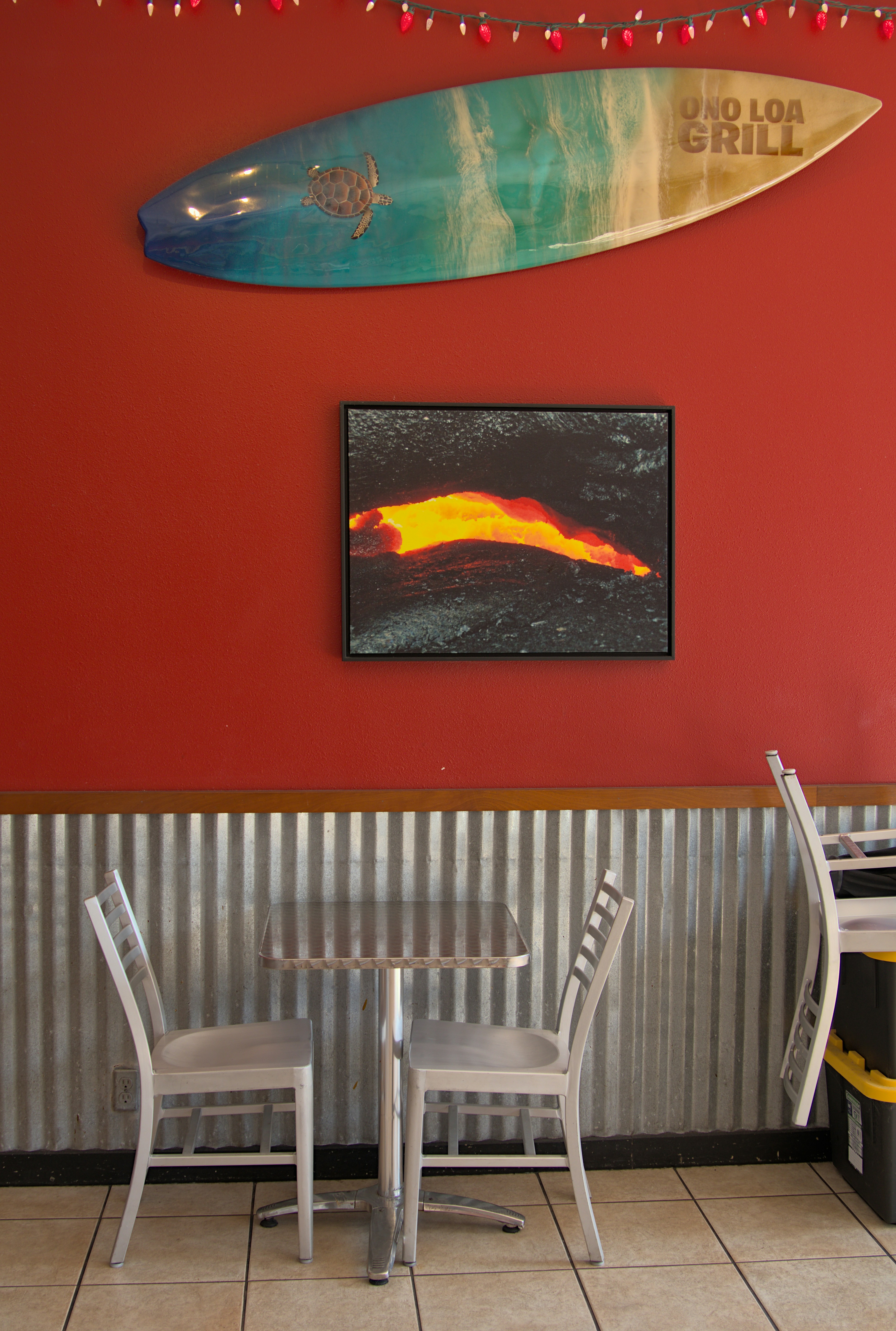 A cozy dining corner featuring a surfboard and a vibrant lava photograph against a red wall, evoking a tropical vibe.