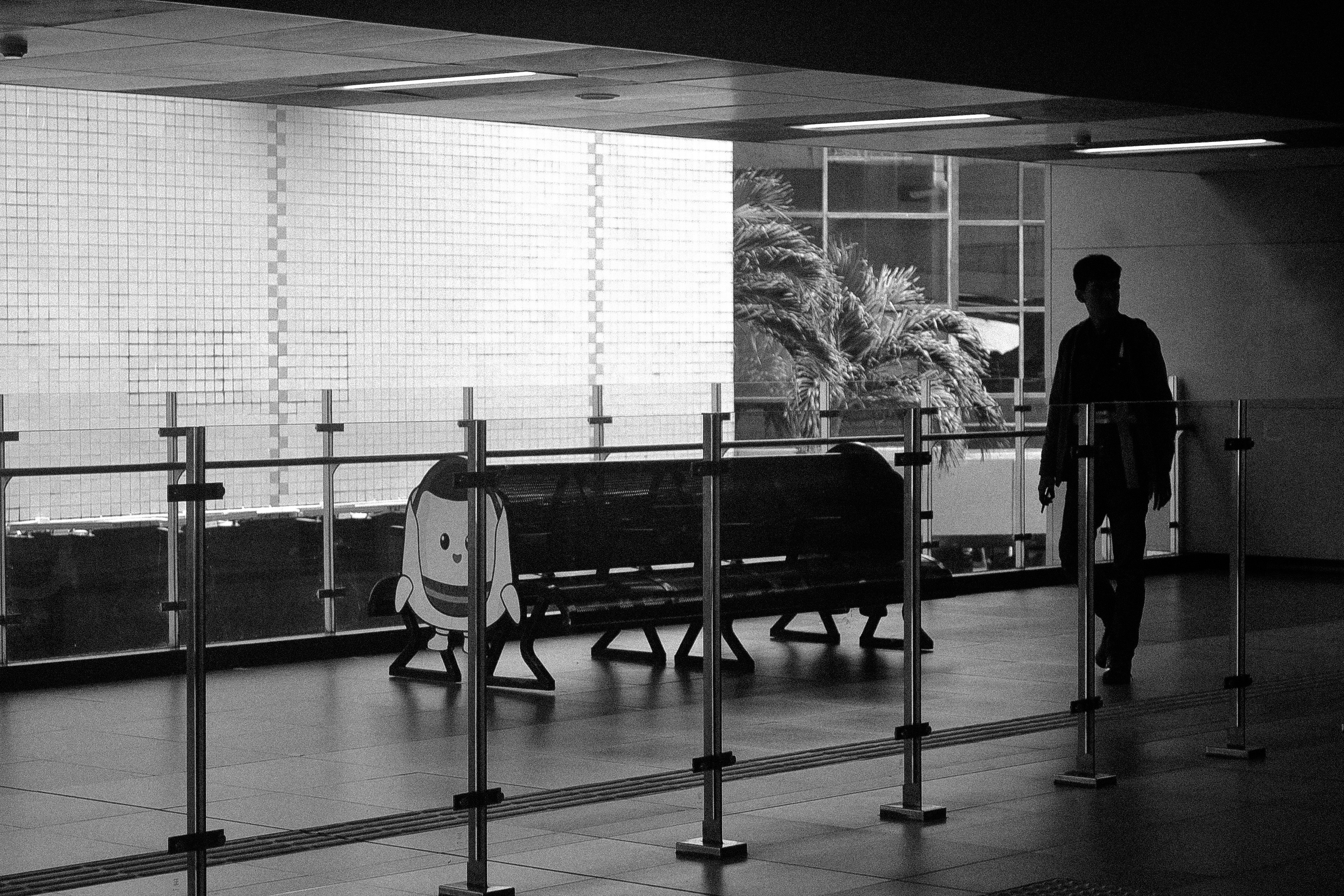 A solitary figure walks past a whimsical bench in a modern transit space, framed by sleek glass barriers and soft natural light.