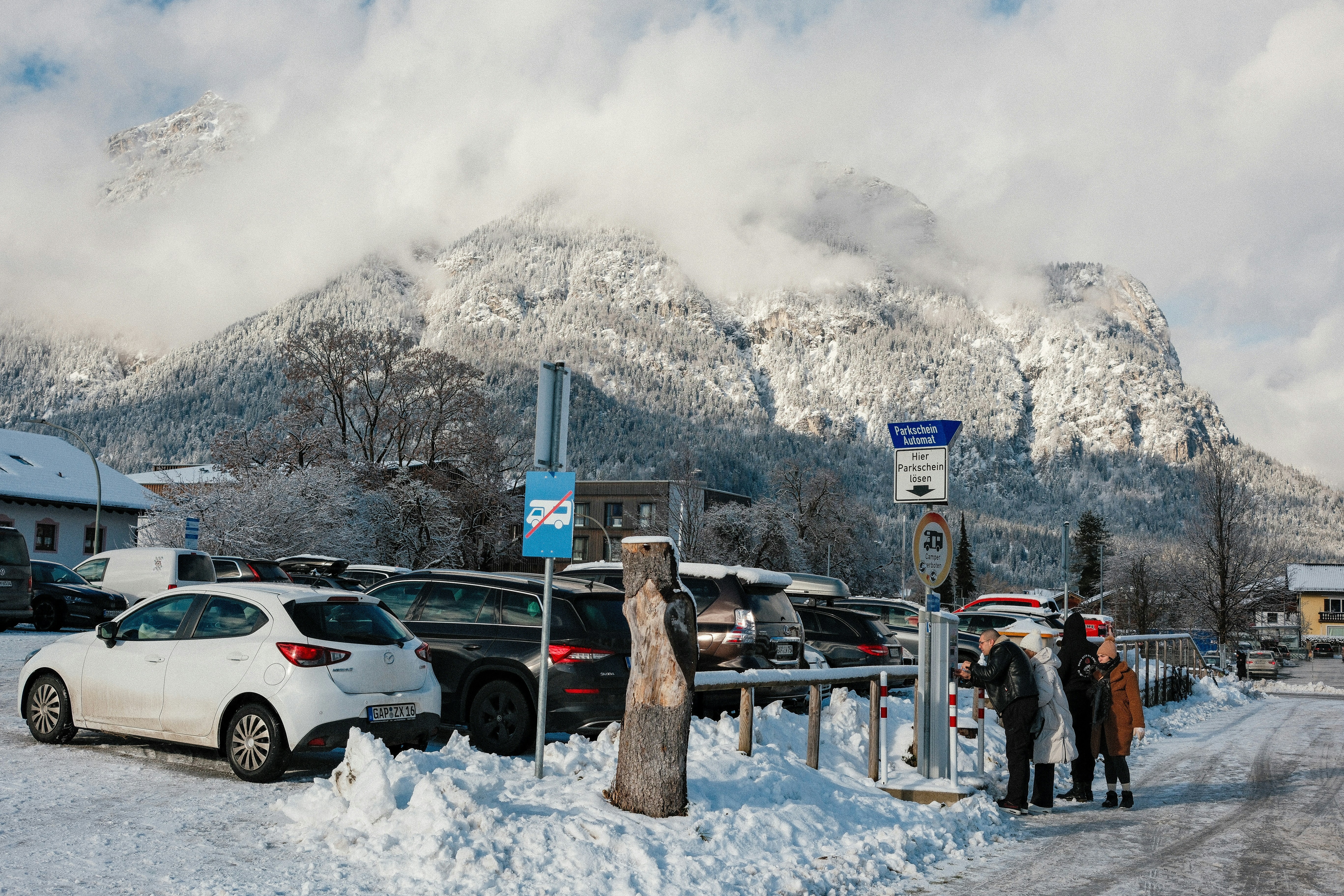 Snow-covered cars and people in a parking lot with majestic mountains shrouded in clouds.