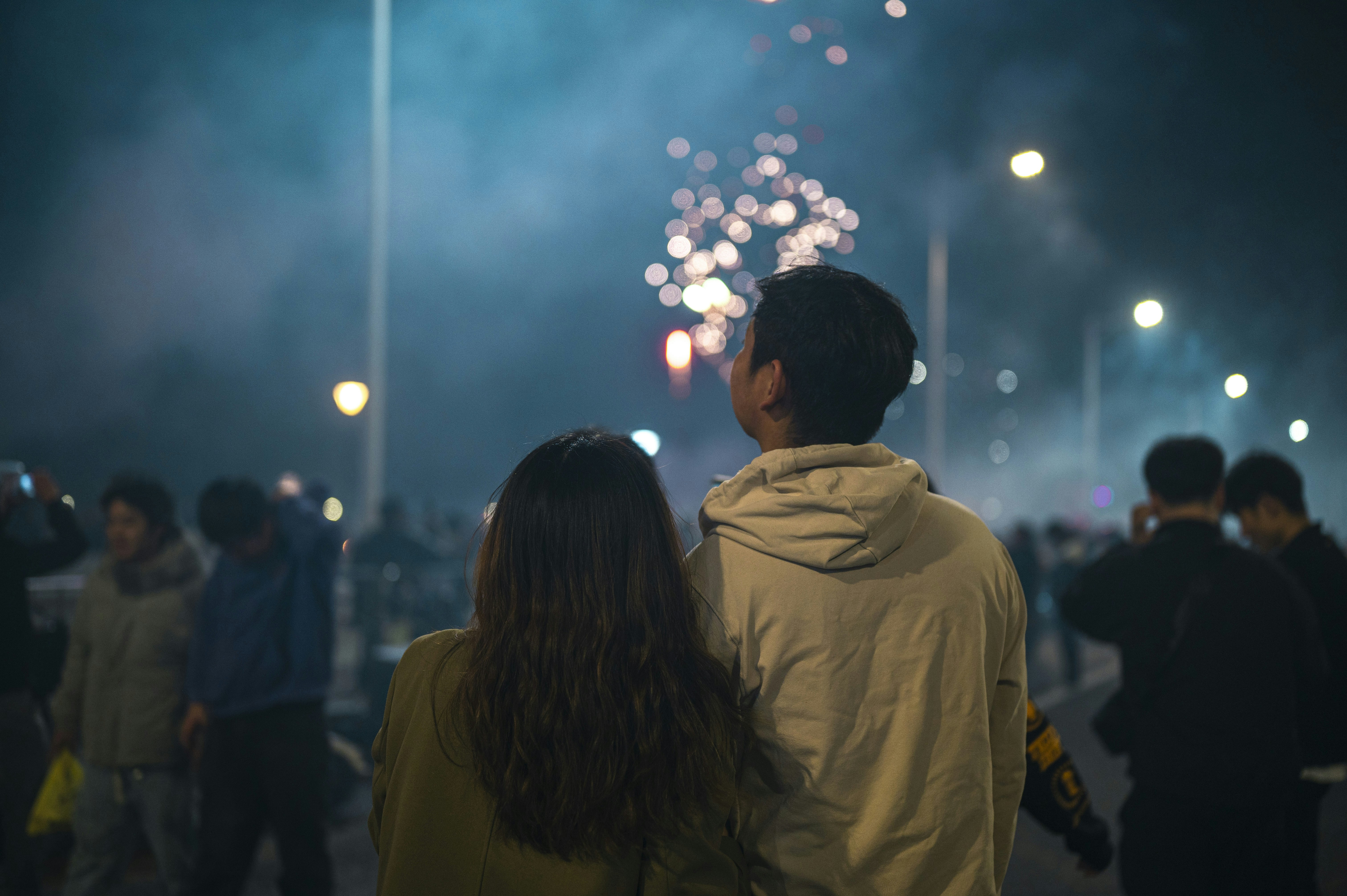 Couple standing together, gazing at fireworks illuminating the night sky amidst a crowd. The atmosphere is filled with excitement and anticipation.