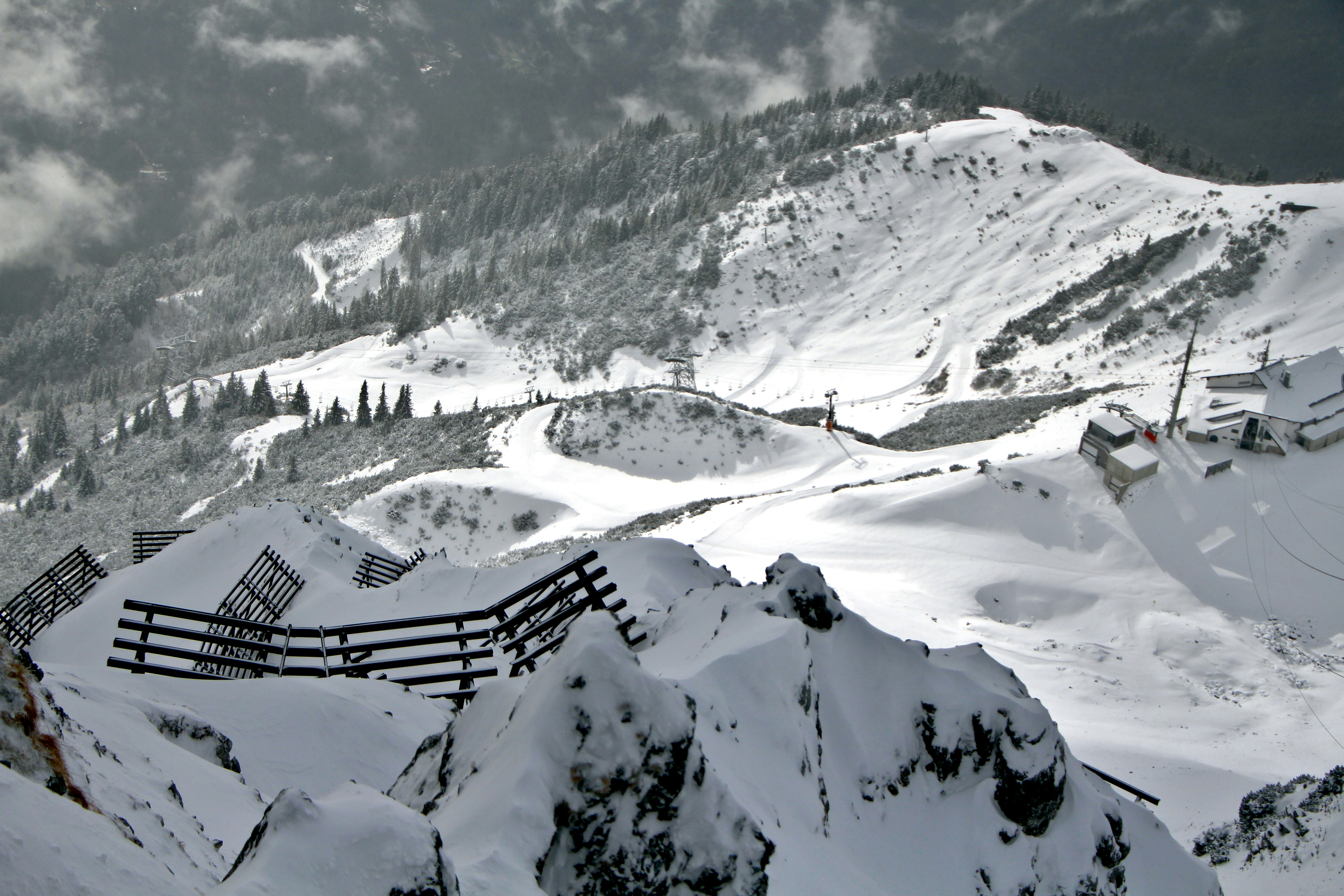 Snow-covered mountain landscape with avalanche barriers and distant forested slopes under a cloudy sky.