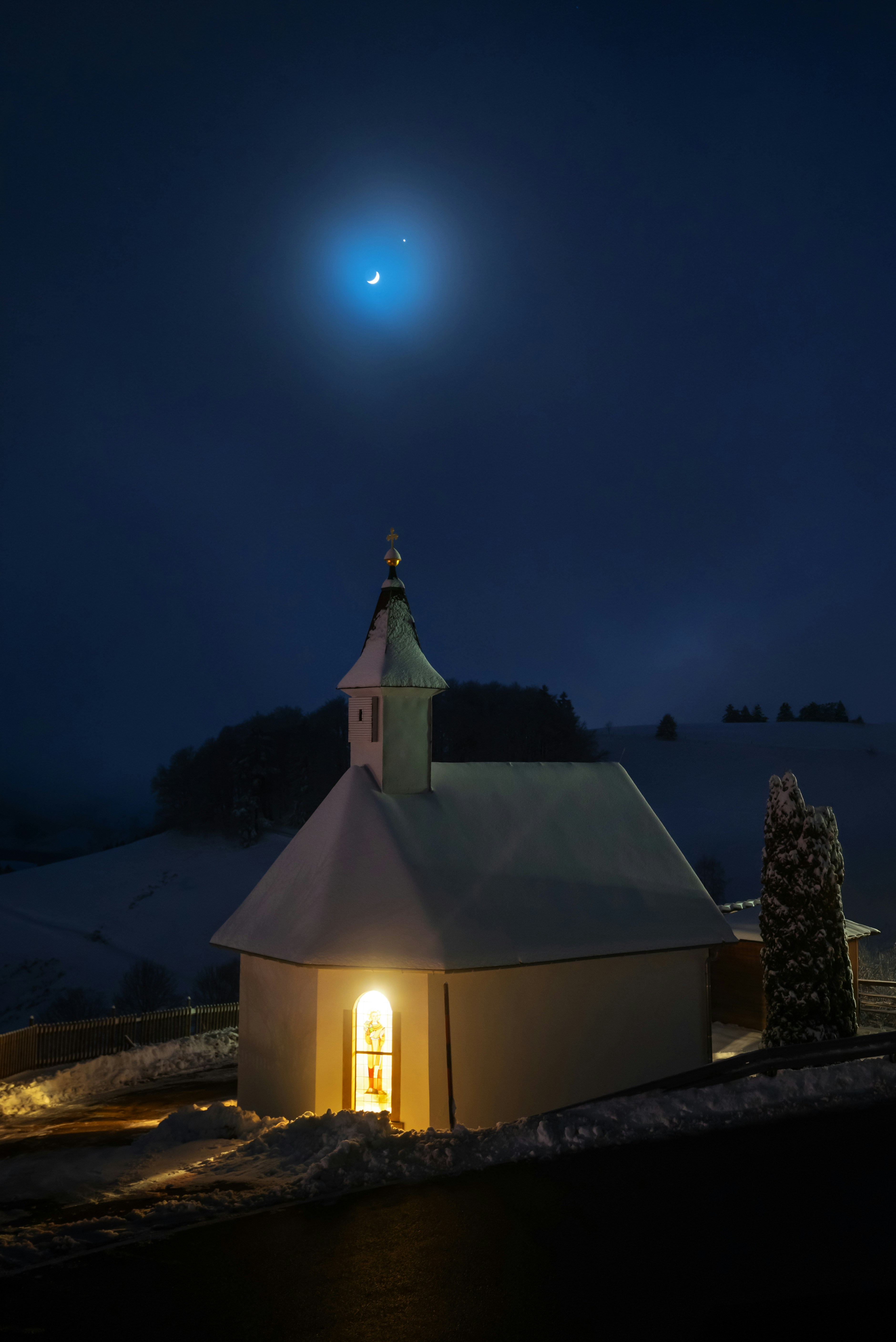 Snow-covered chapel softly lit from within under a crescent moon in a deep blue sky.