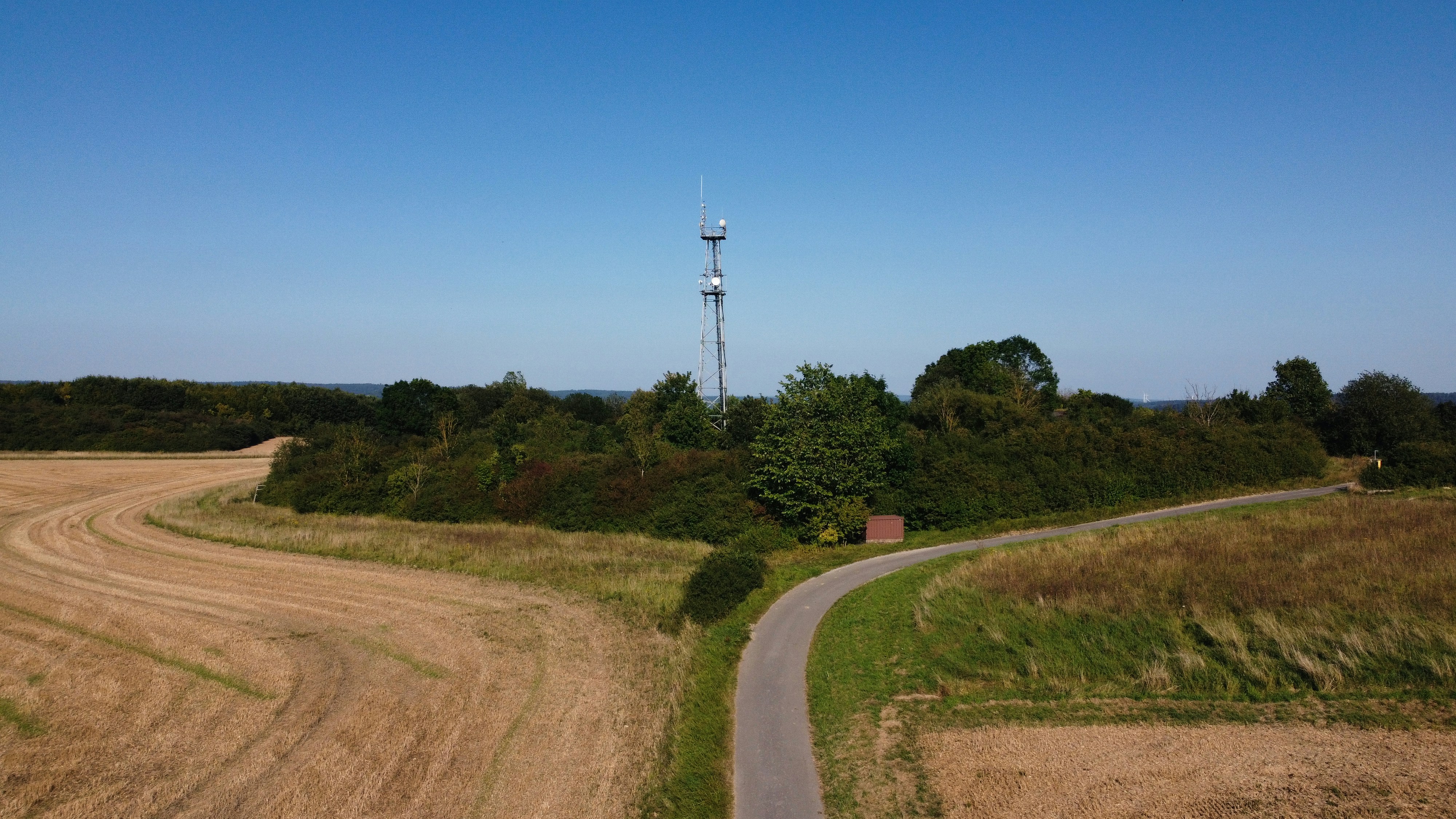 Aerial drone photograph of a rural landscape featuring a winding road through fields, with a tall communications tower and scattered trees under a clear blue sky.