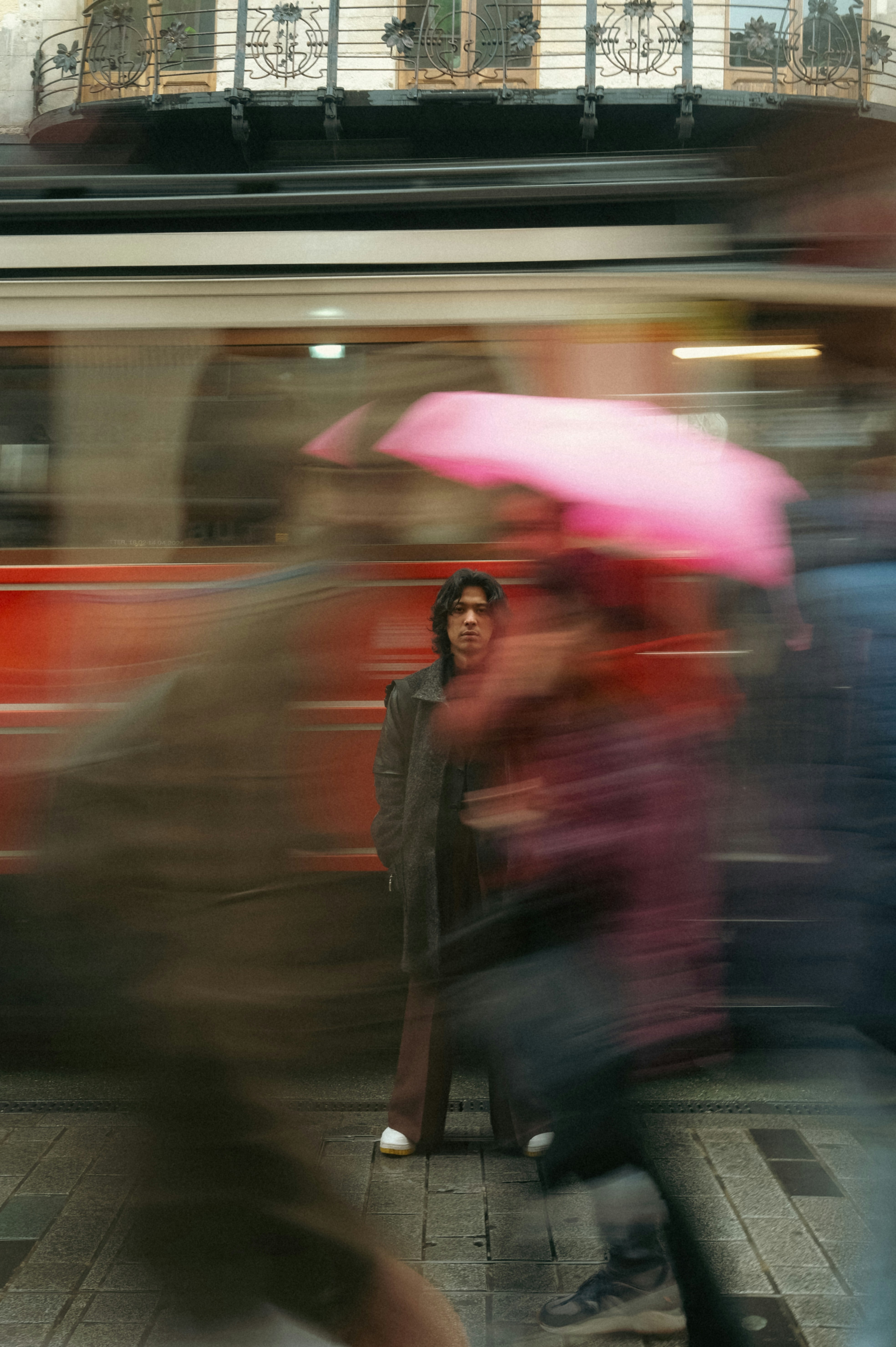 Figure standing still on a bustling street, surrounded by blurred motion of people with umbrellas. Vibrant red and pink hues contrast the muted cityscape.