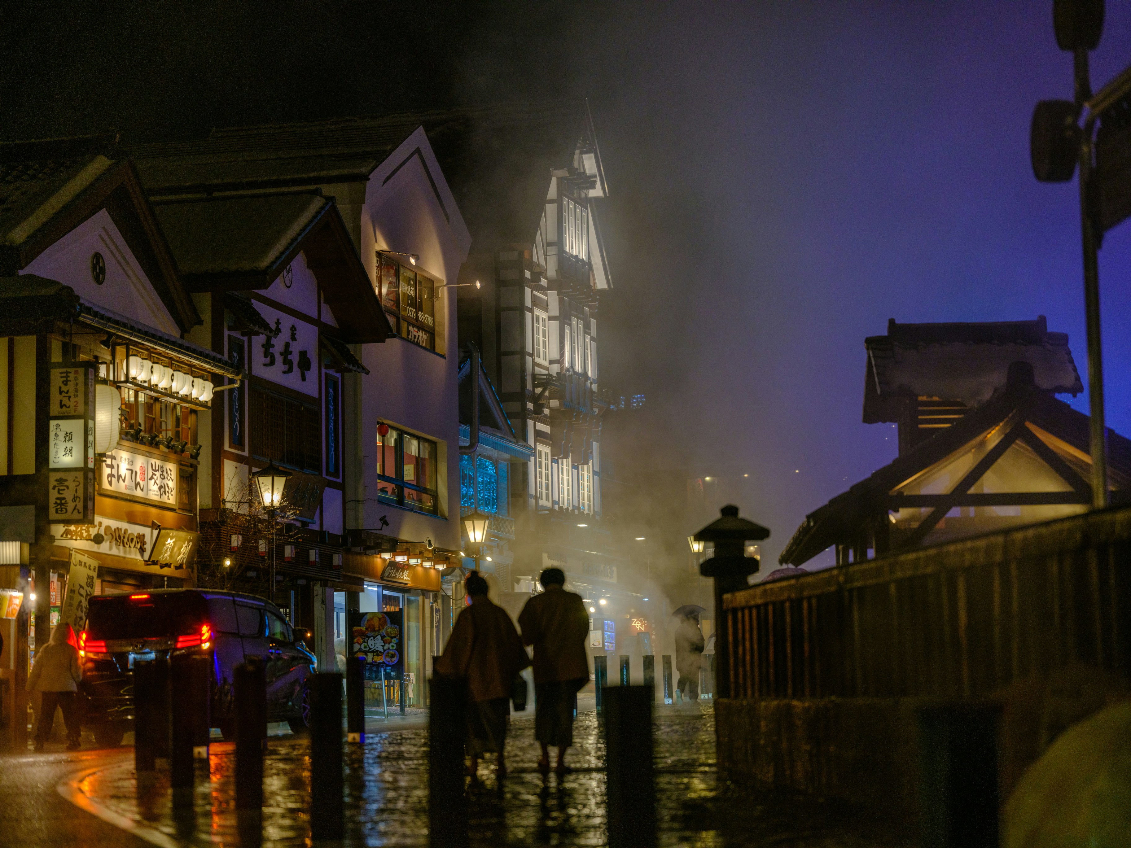 Silhouetted figures walk along a misty street lined with warmly lit buildings at night.