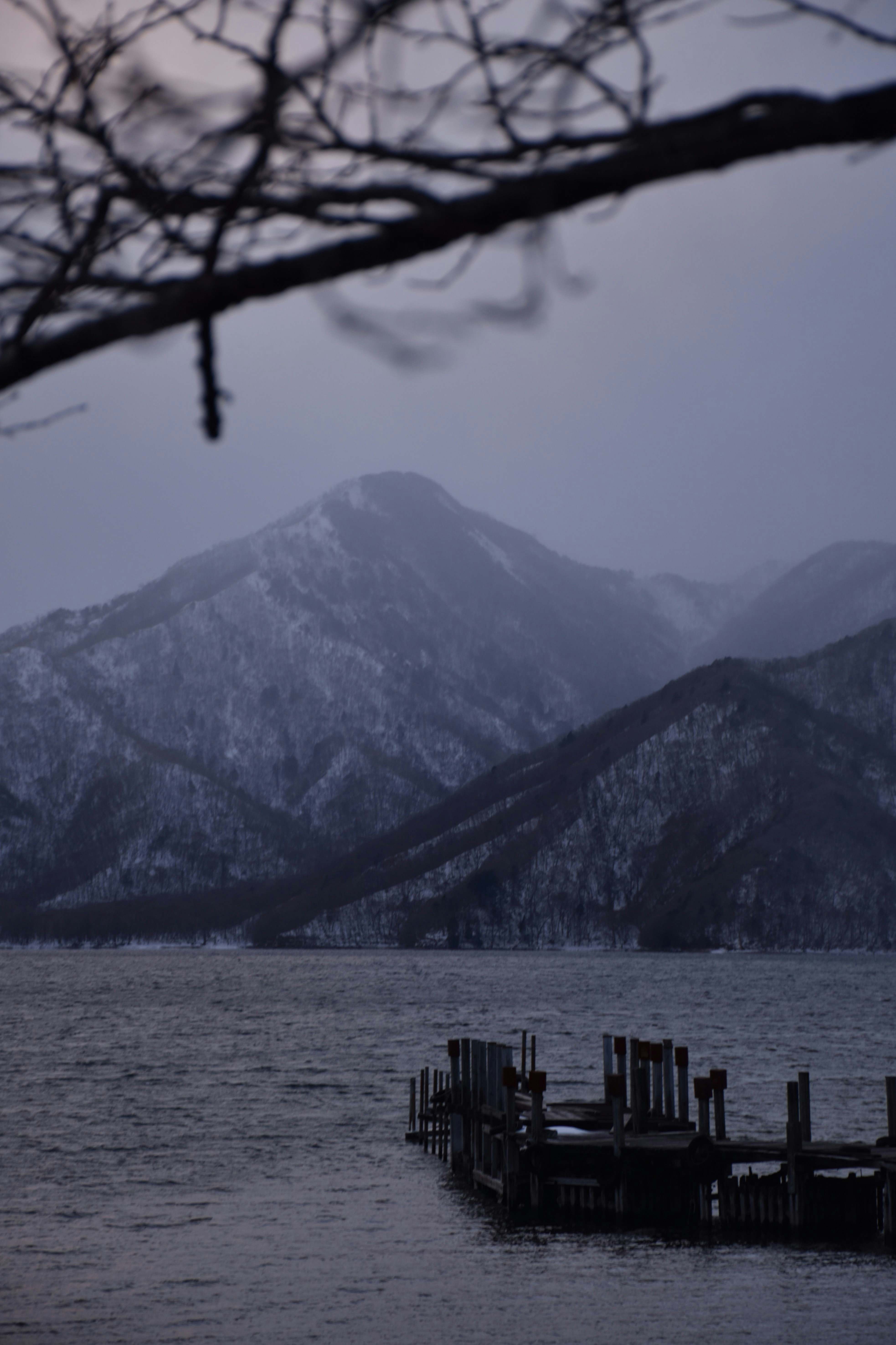 A weathered dock extends into a tranquil, icy lake, framed by snow-capped mountains under a muted sky.