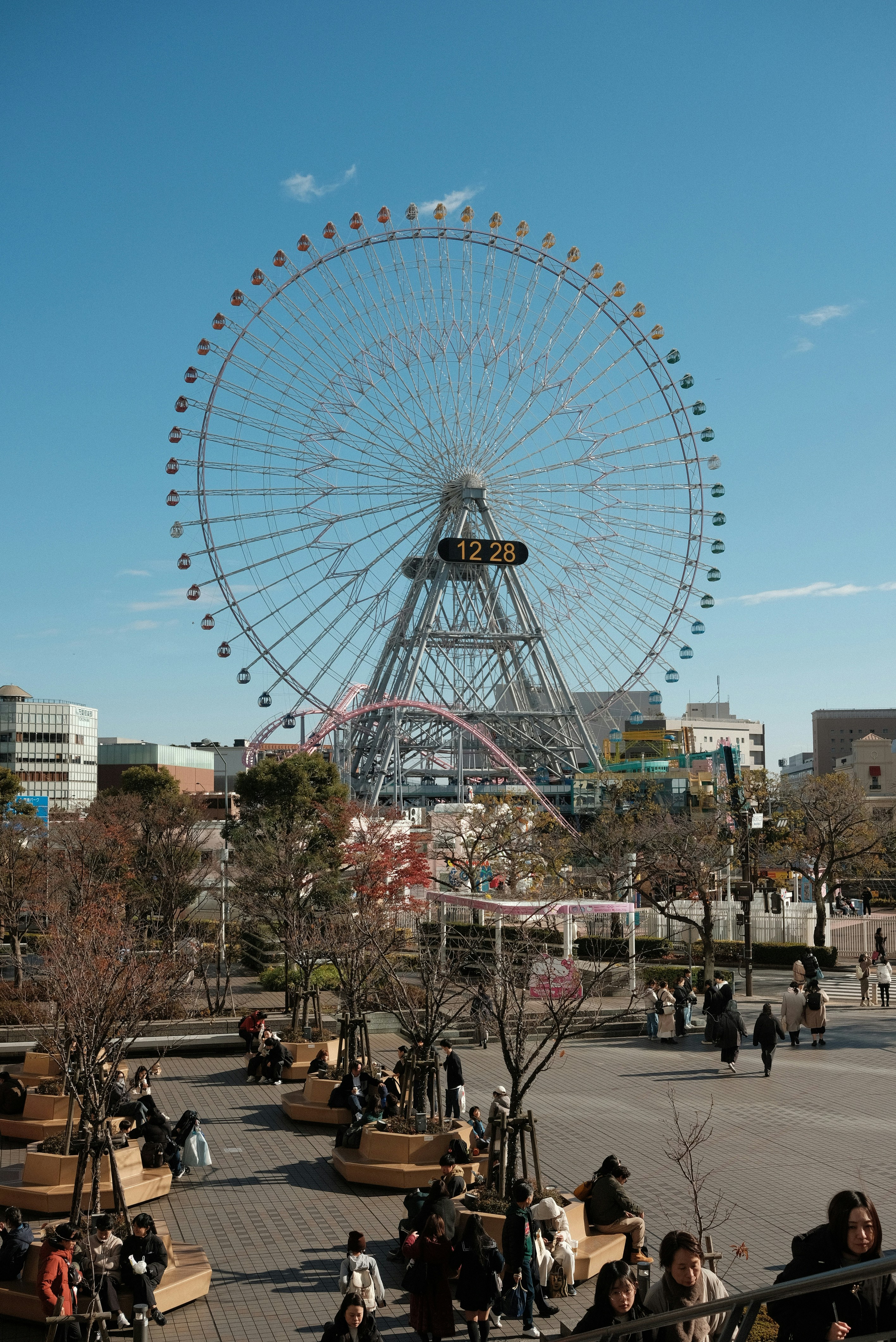 Ferris wheel towering over a bustling fairground, with visitors enjoying leisure time below. The clock shows 12:28.