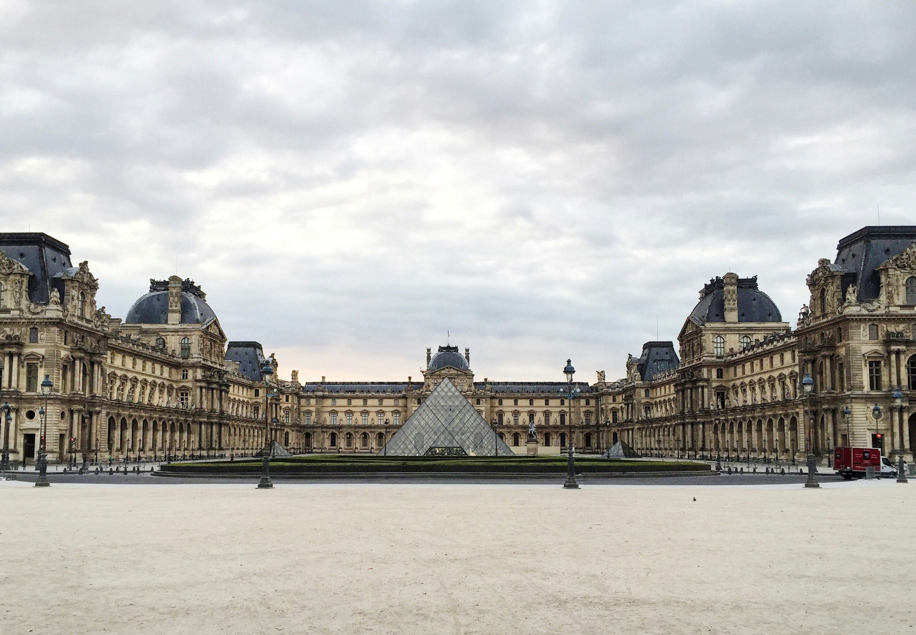 The Louvre Museum's glass pyramid stands amidst historic architecture under a cloudy sky.
