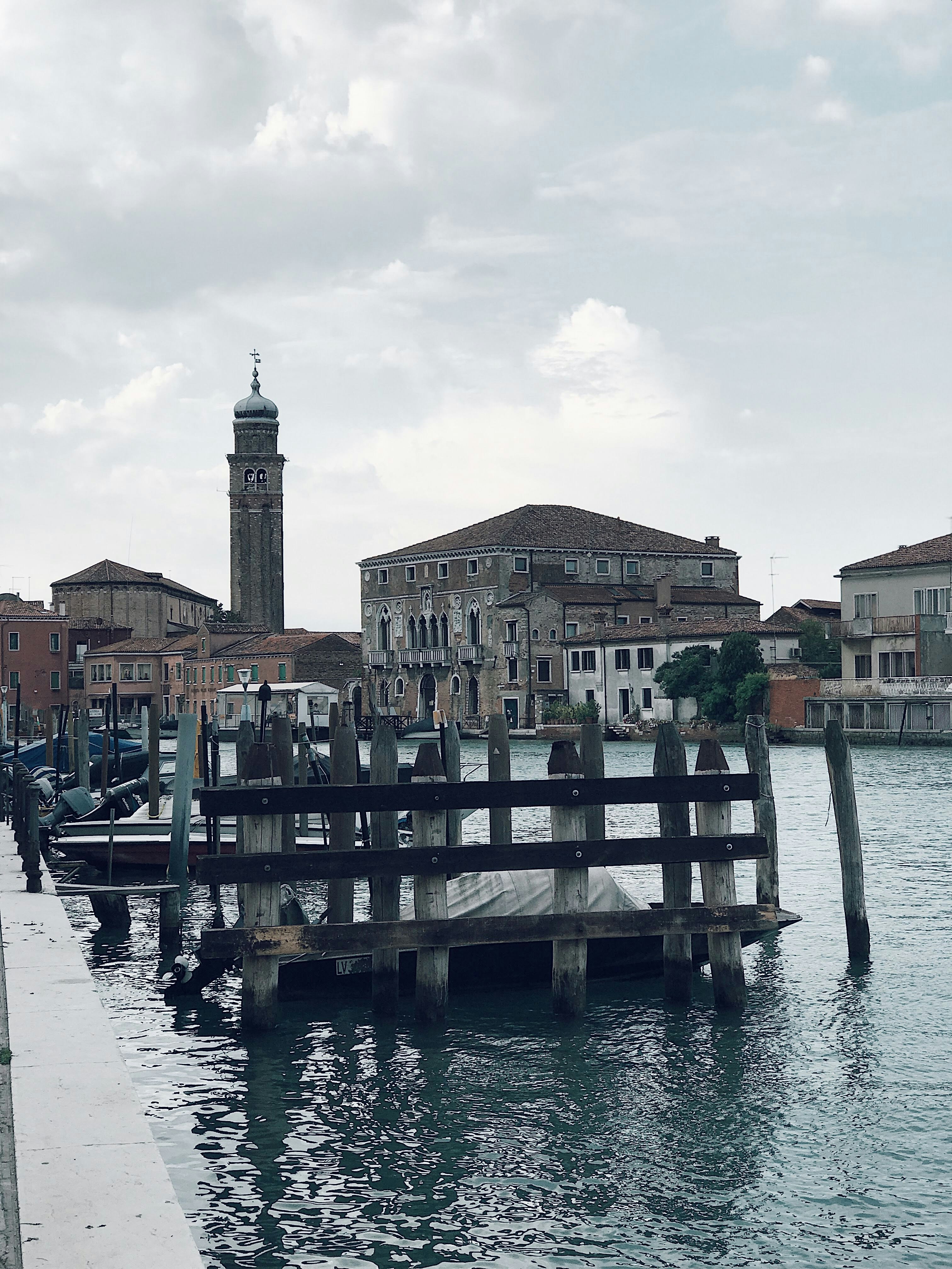 Wooden dock extending into a calm canal with historic buildings and a tall bell tower in the background under a cloudy sky.