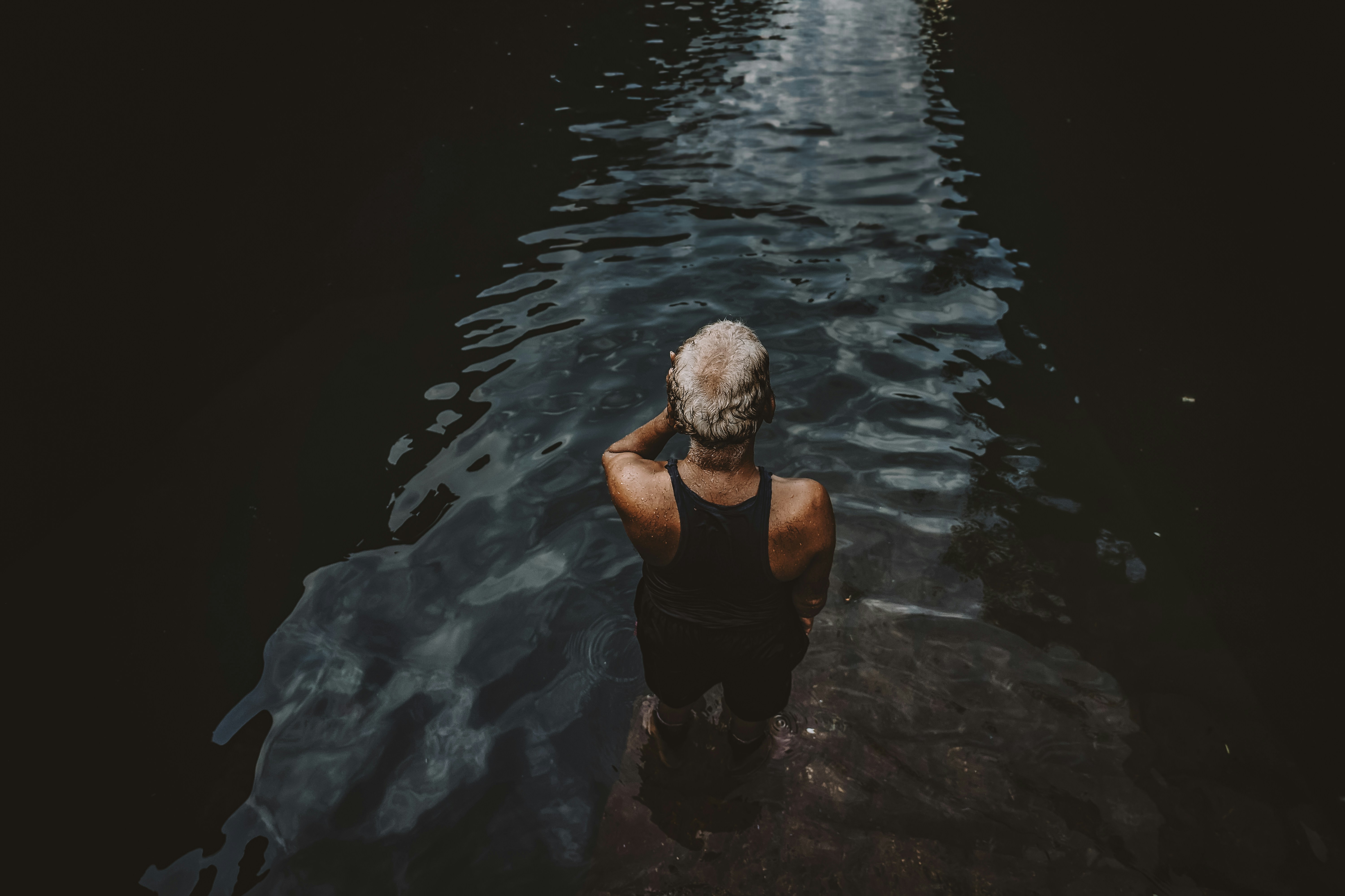A person stands knee-deep in dark water, gazing into the distance, surrounded by shadowy tranquility. The rippling surface reflects a hint of light above.