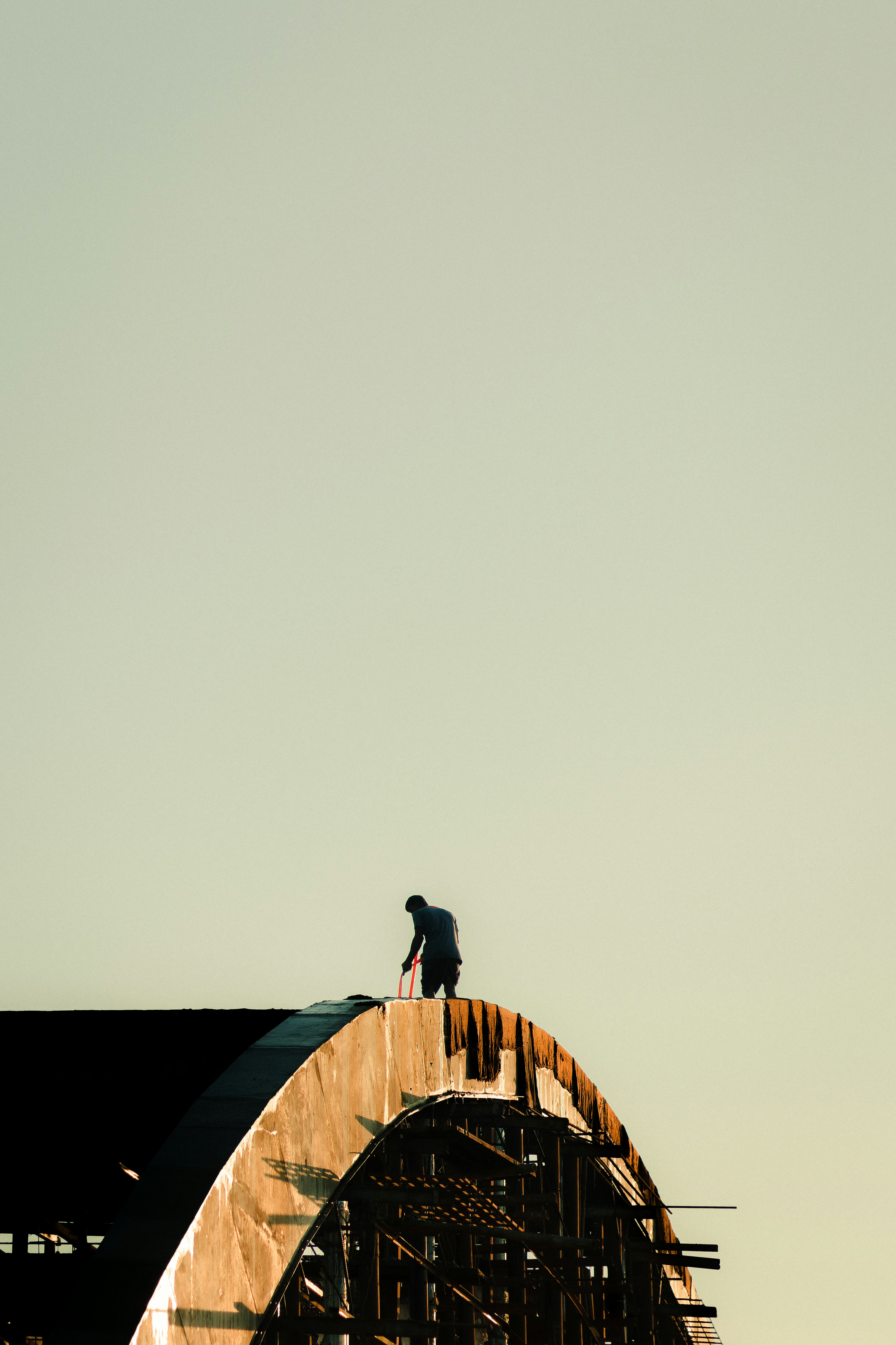 Edgewalker on the Curved Shell: A lone silhouette atop rusted architecture against a vast pale sky
