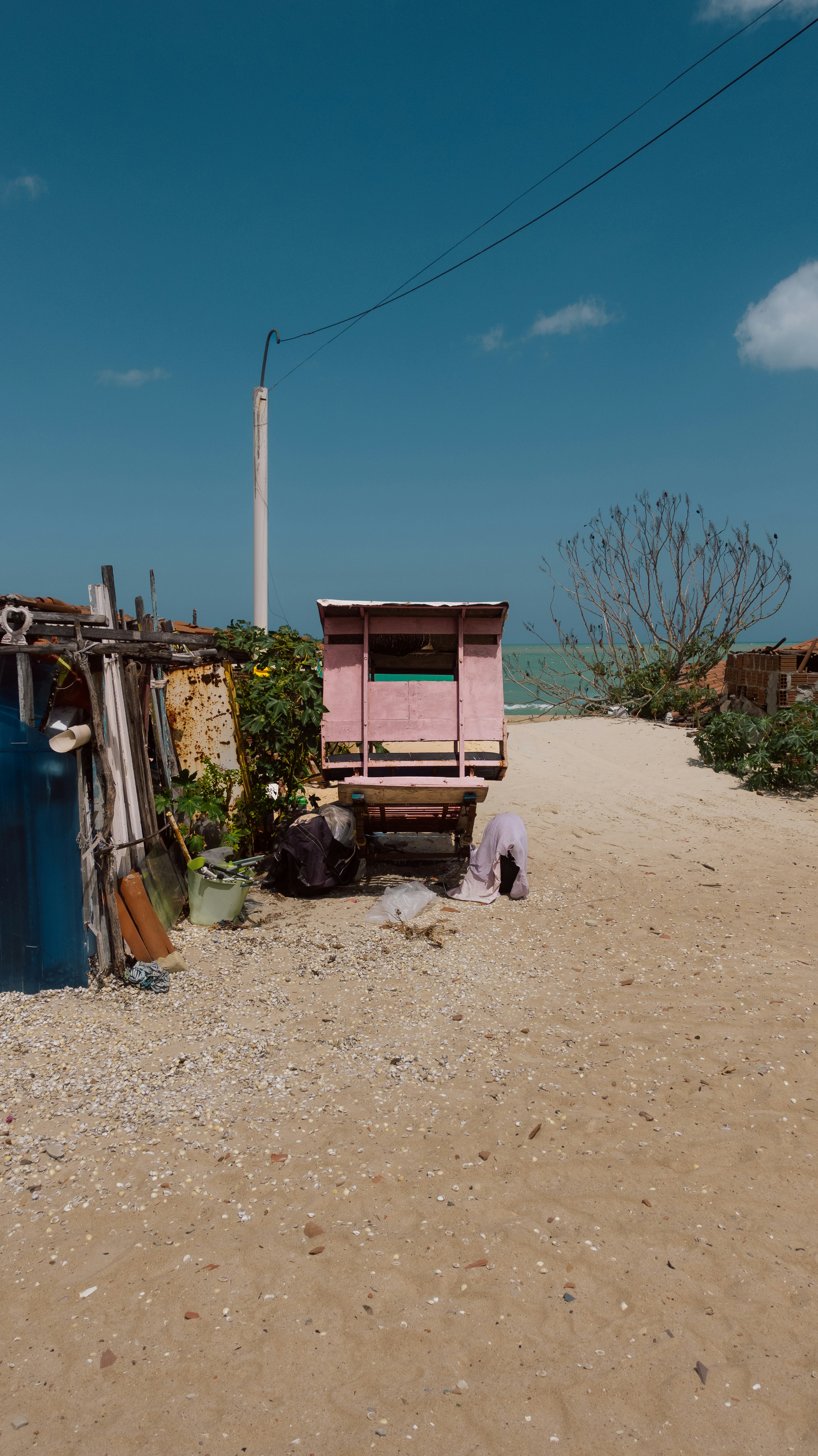 Vibrant pink hut on a sandy beach with weathered structures to the left and calm turquoise sea in the background.