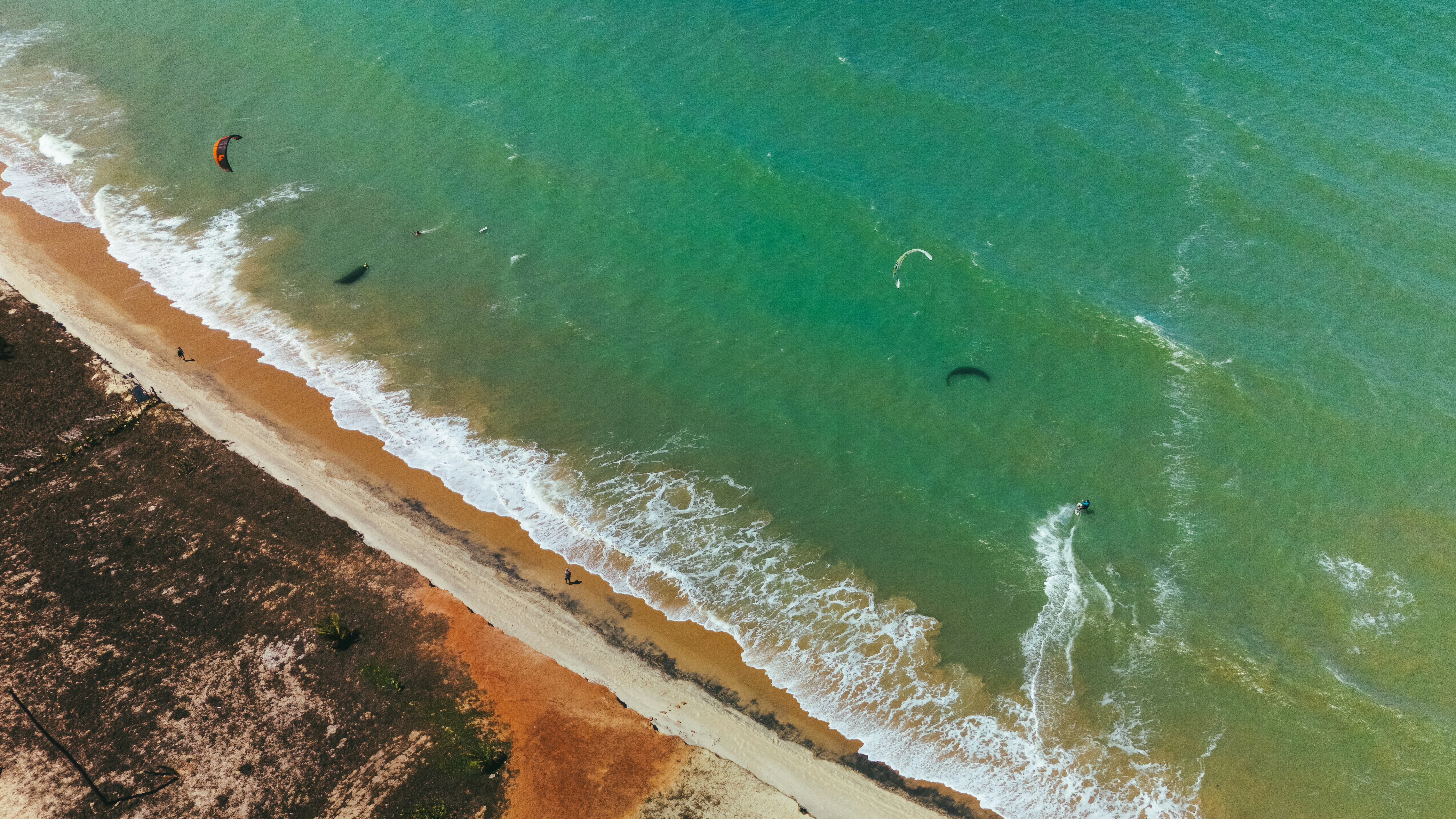 Aerial view of kitesurfers navigating turquoise waves along a sandy shoreline.
