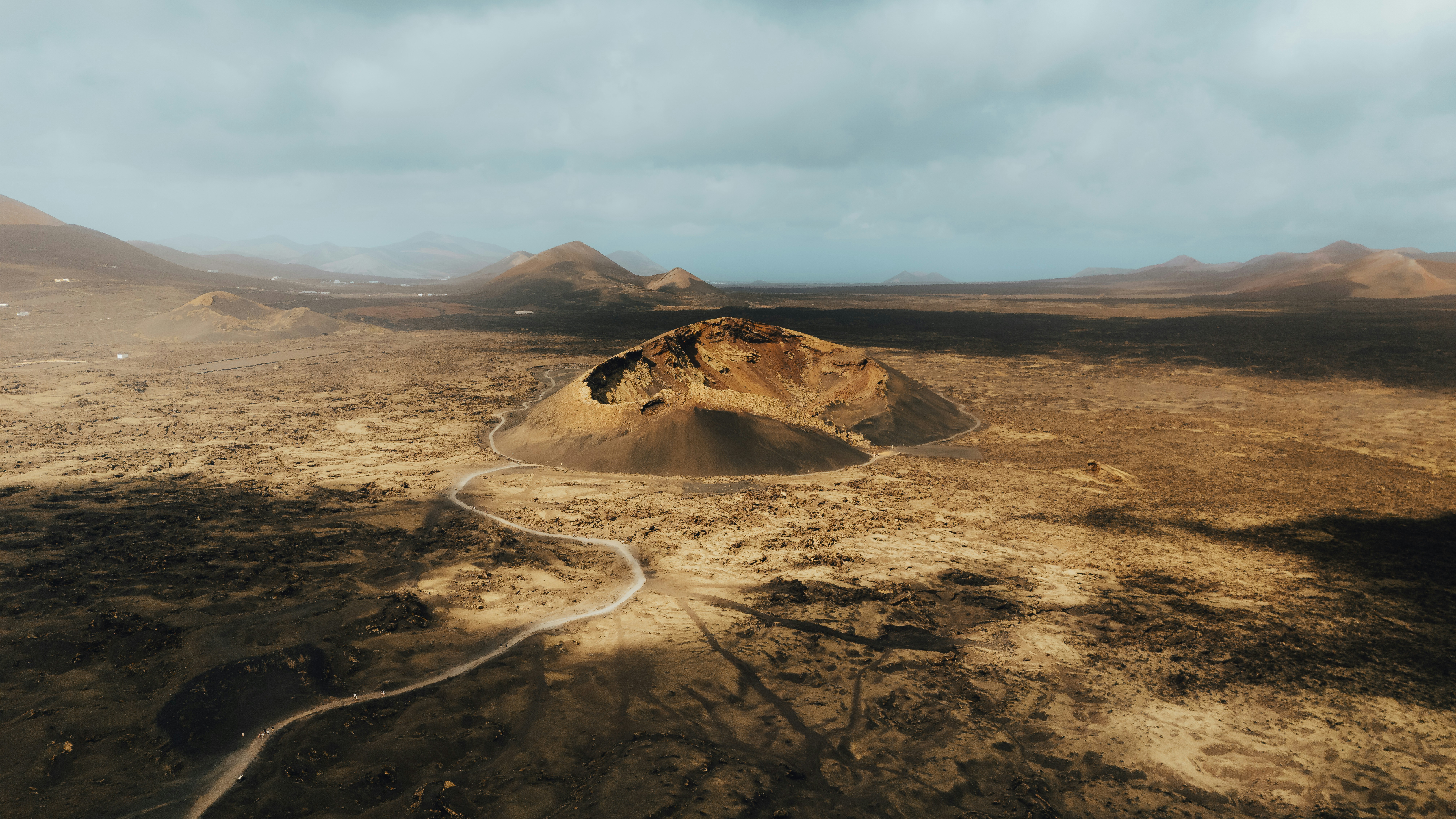 Aerial view of a rugged volcanic terrain with a prominent crater and winding road under a cloudy sky.