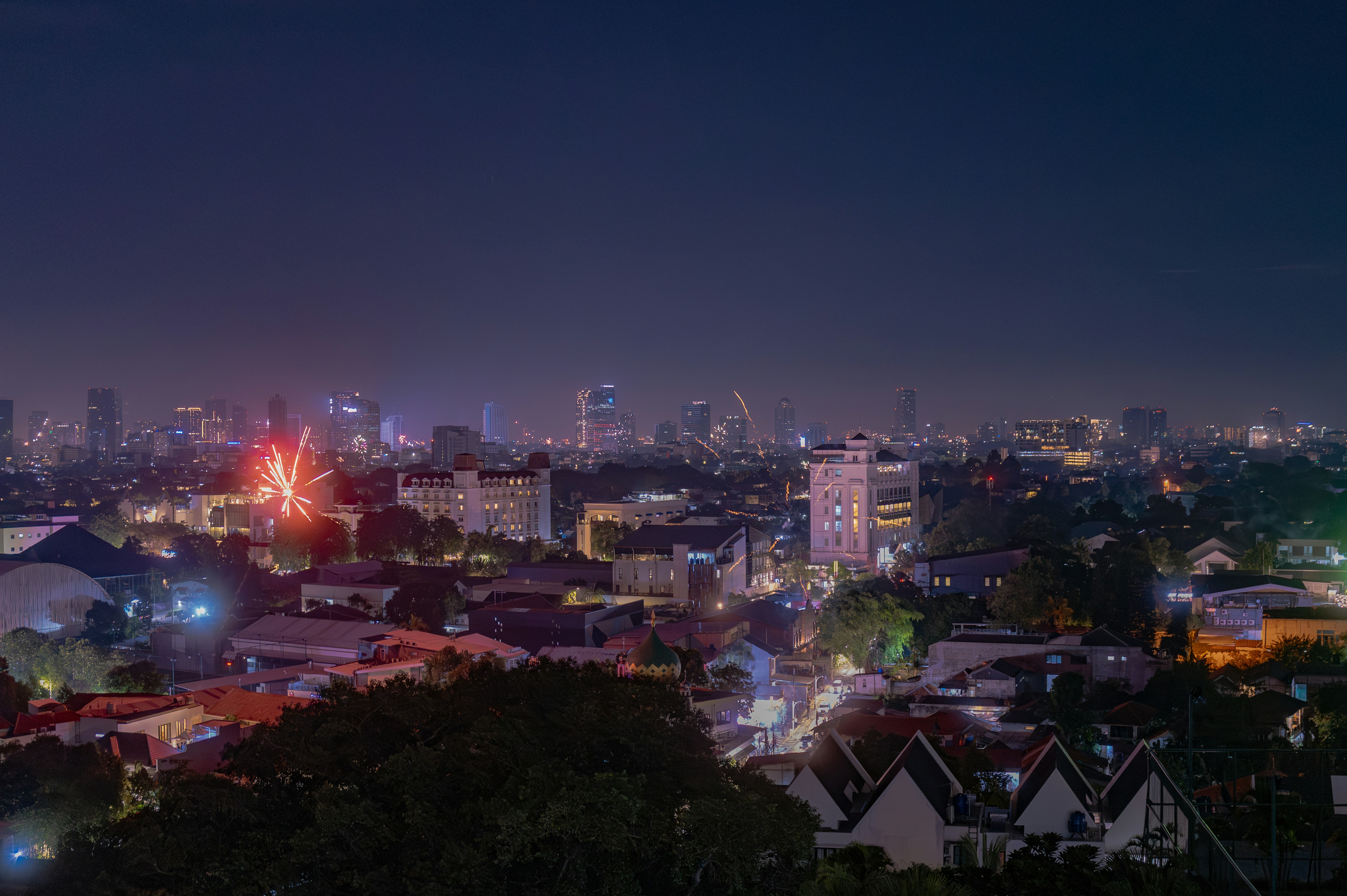 Night cityscape with a crimson firework bursting over a suburban foreground. Distant city lights create a moody, atmospheric backdrop.