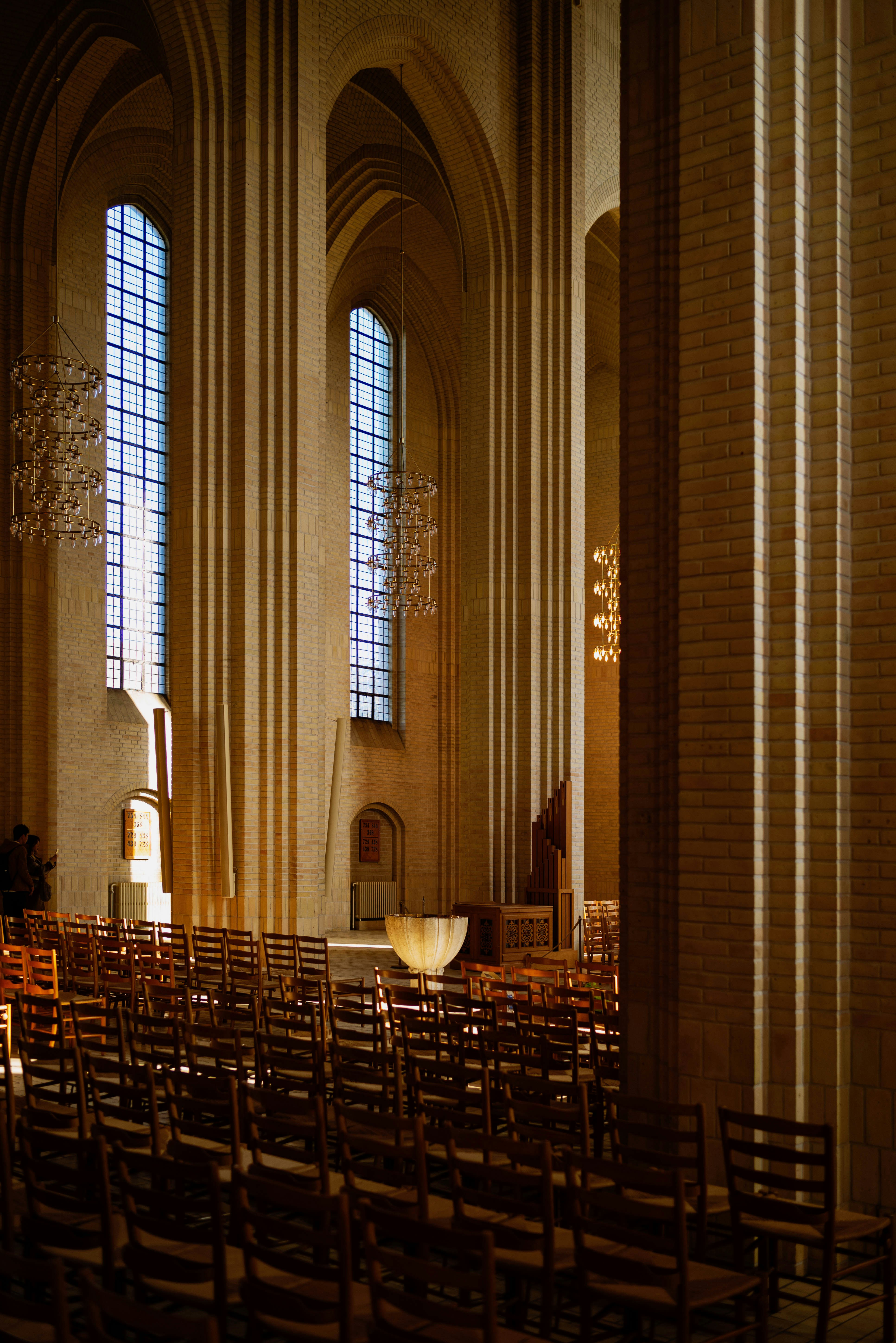 Wooden chairs in rows beneath soaring arches and stained glass, bathed in warm light, showcasing intricate brickwork.
