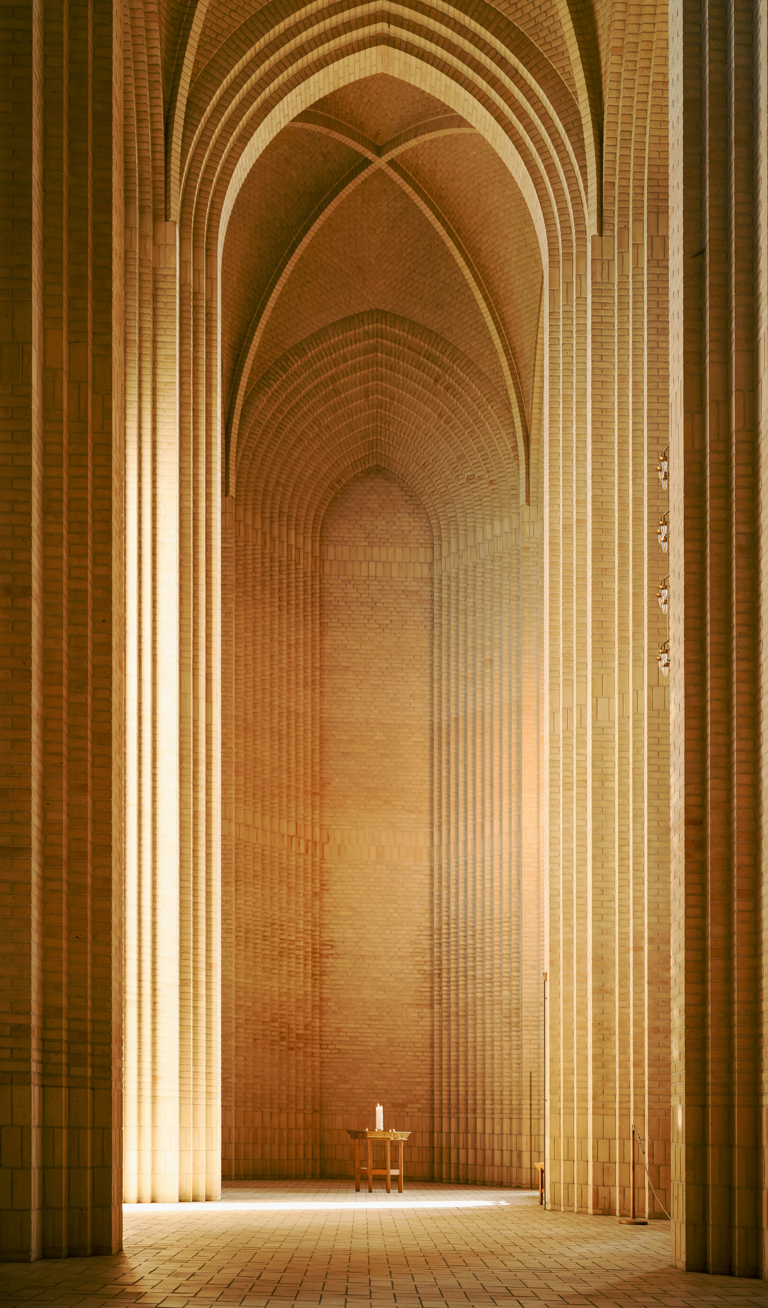 Grand hallway with soaring arches and intricate brickwork illuminated by warm light. A solitary bench sits at the center, enhancing the vertical rhythm of towering pillars.