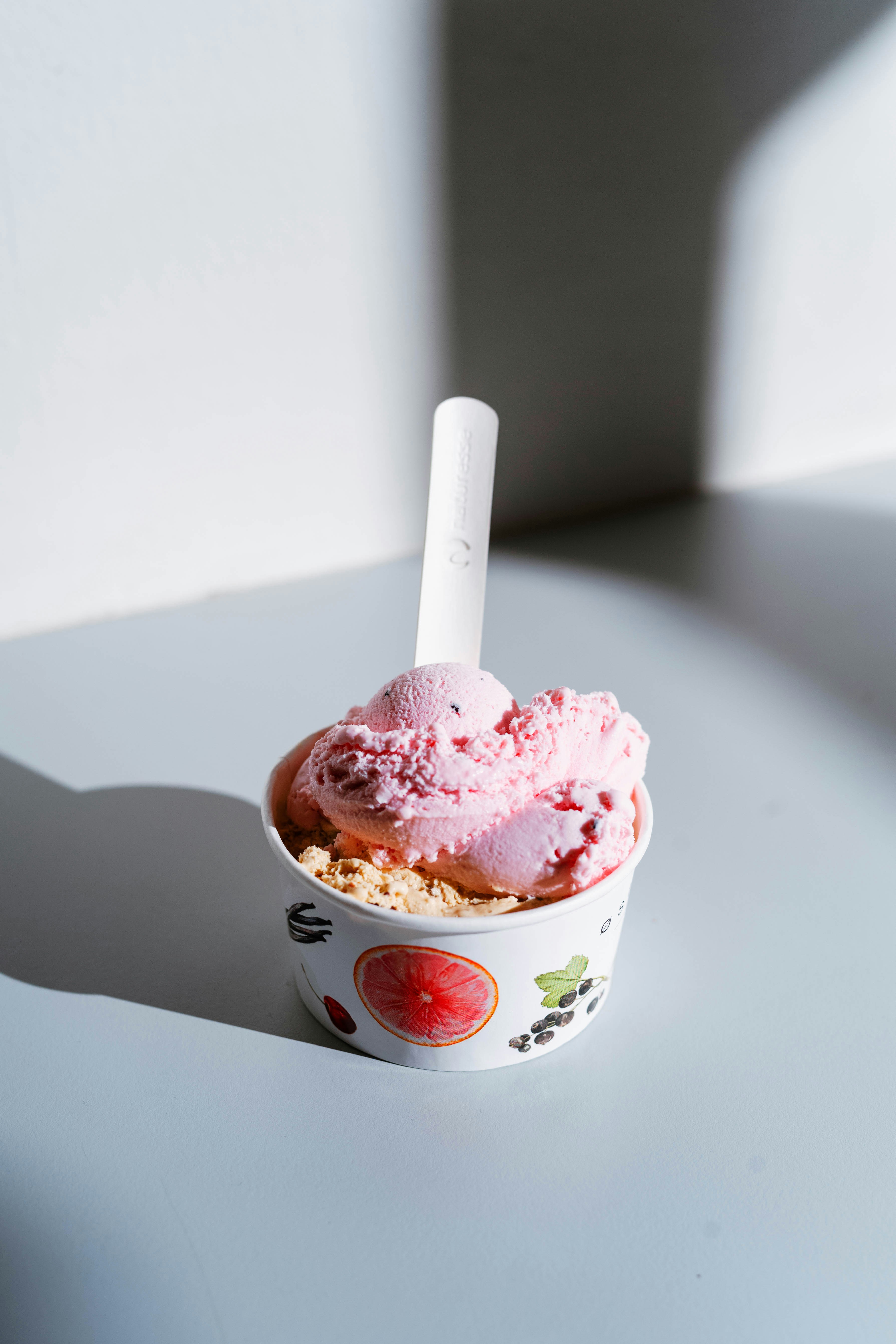 Pink ice cream scoops crown a small patterned cup on a pale white table, with sharp diagonal shadows.