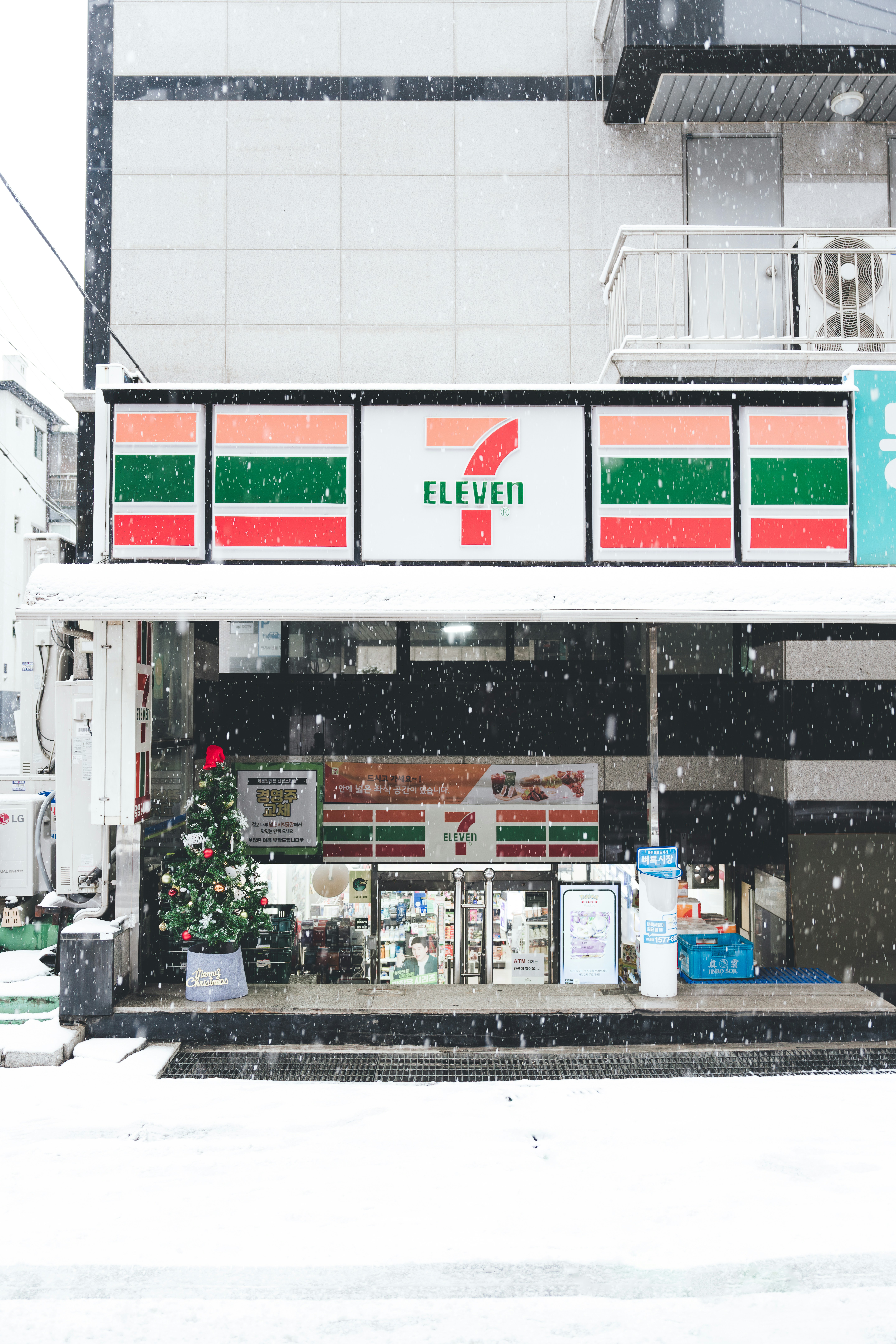 7-Eleven storefront in snowfall with a decorated Christmas tree and vibrant signage.