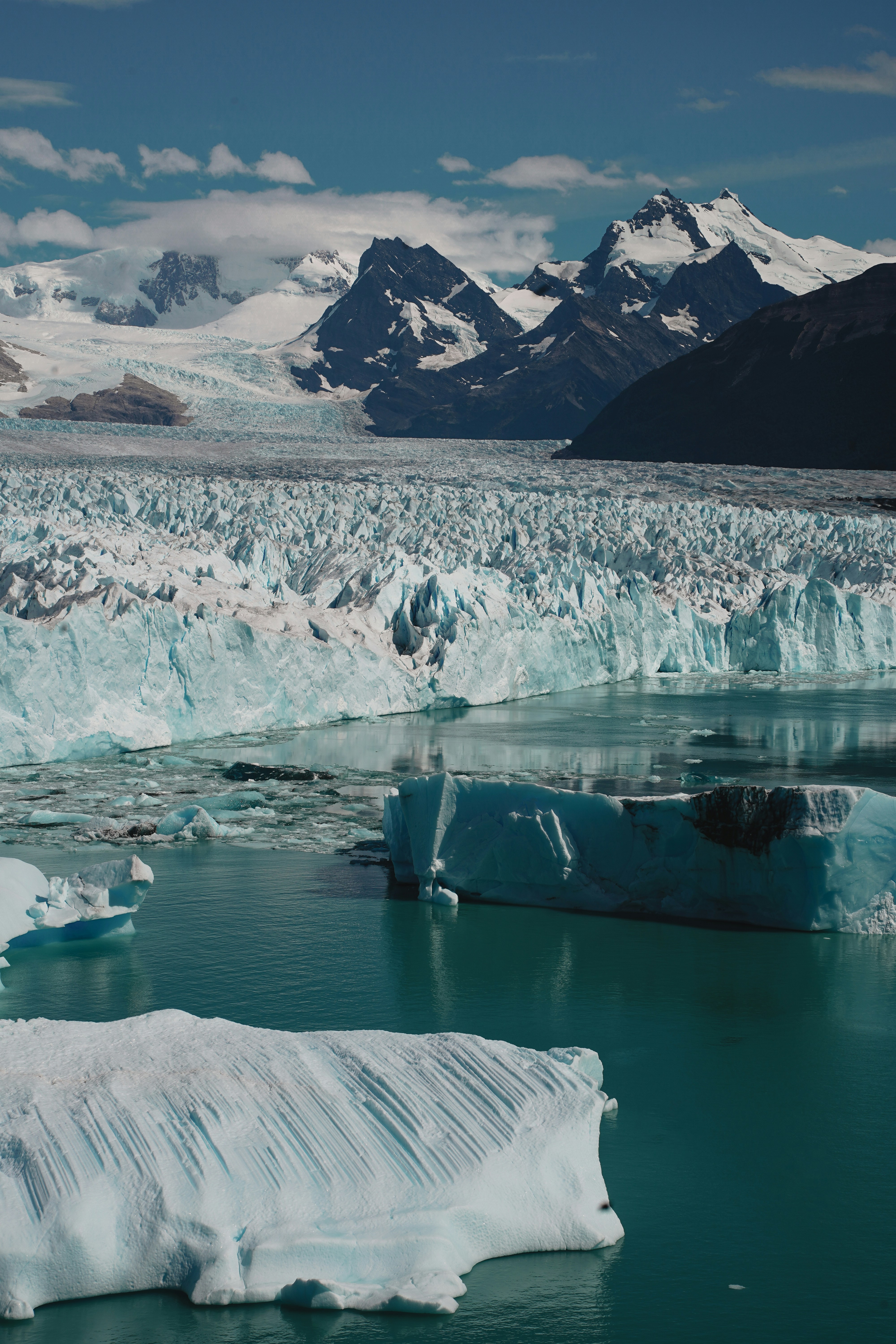 Vast icebergs float in a tranquil glacial lagoon, framed by towering mountains under a clear blue sky.