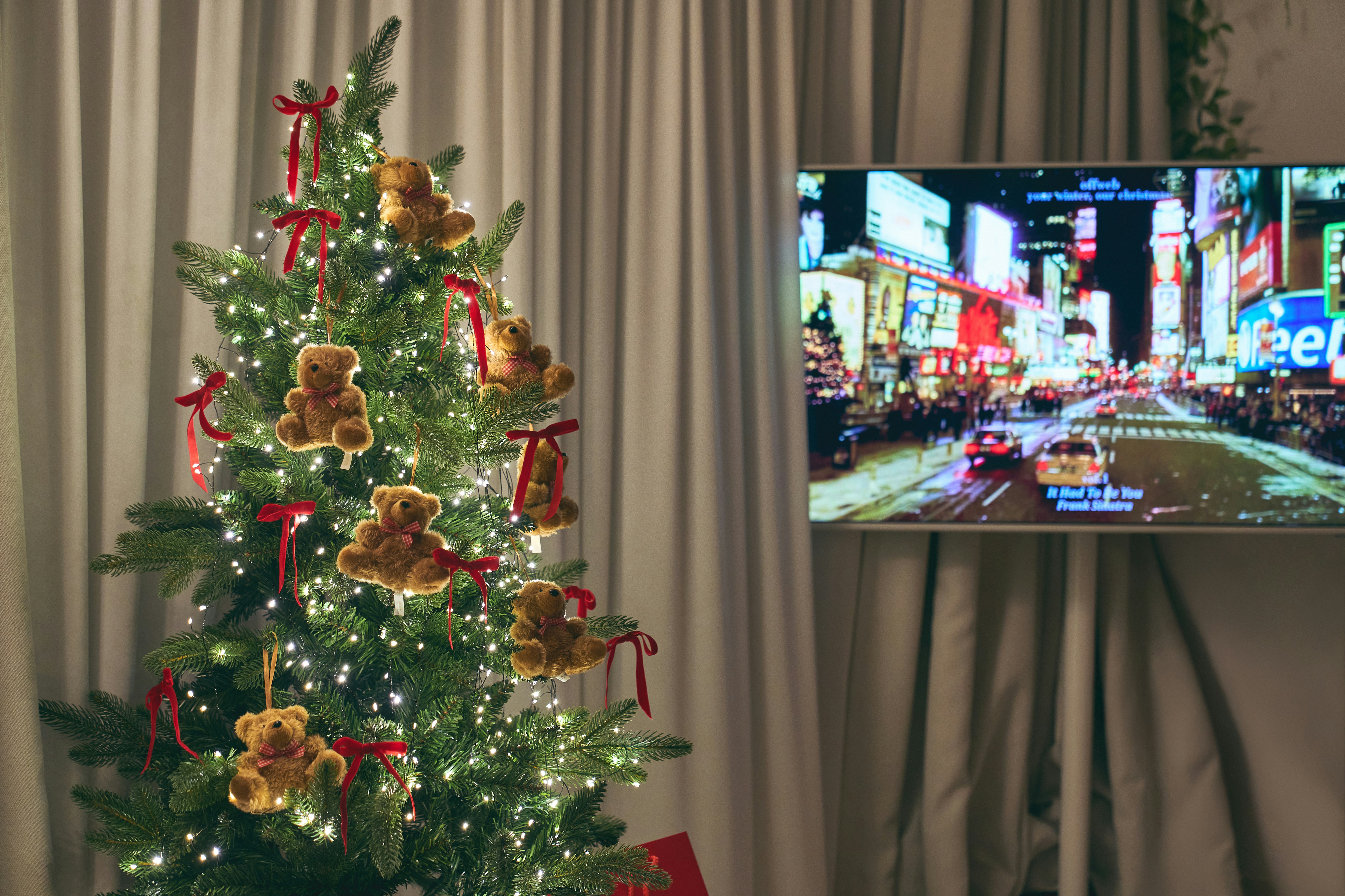 Christmas tree adorned with teddy bear ornaments and ribbons, illuminated by fairy lights, beside a television displaying a vibrant city scene.