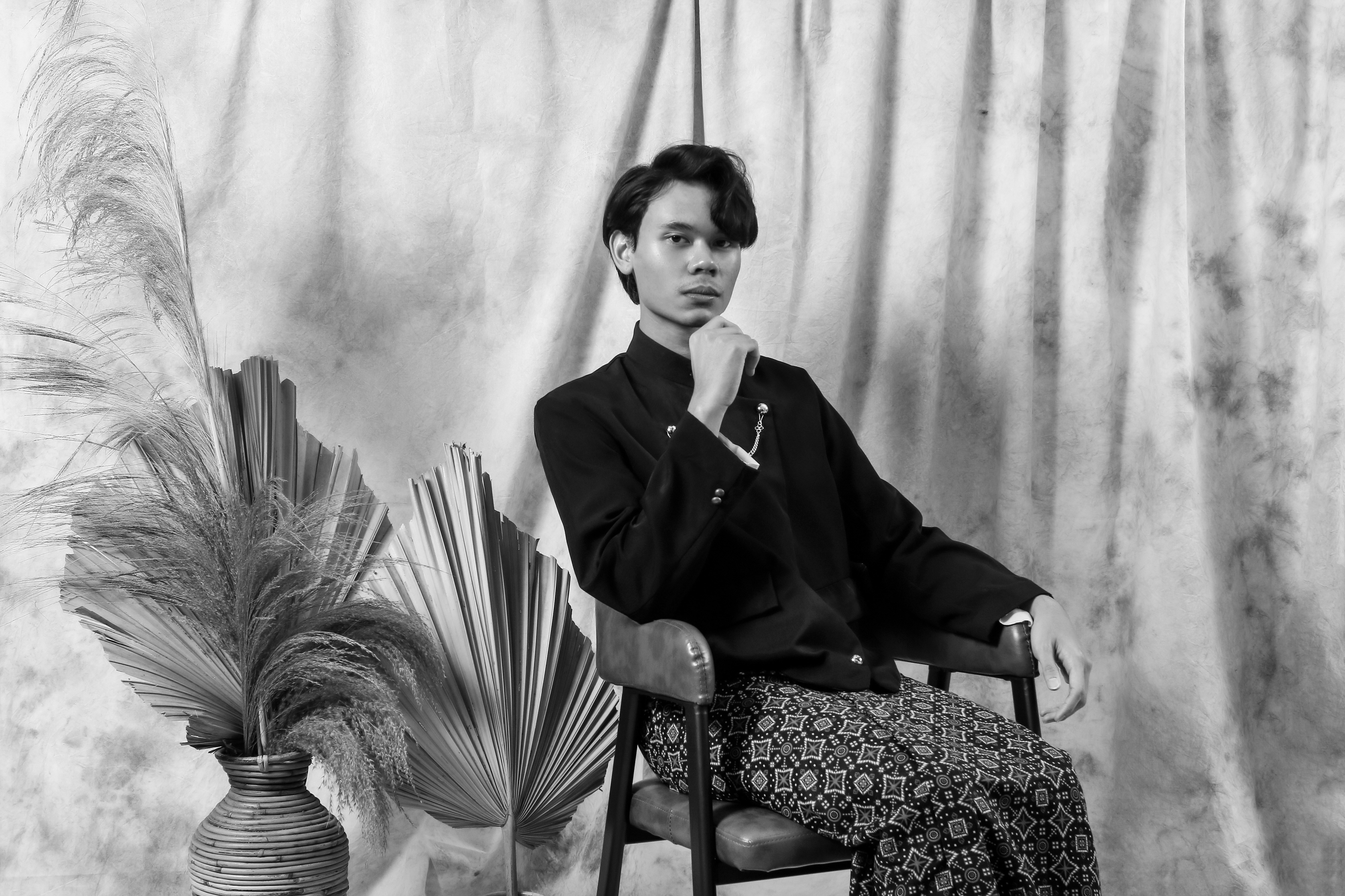 Person seated in a chair wearing traditional attire with dried palm leaves in a vase, set against a textured draped background.