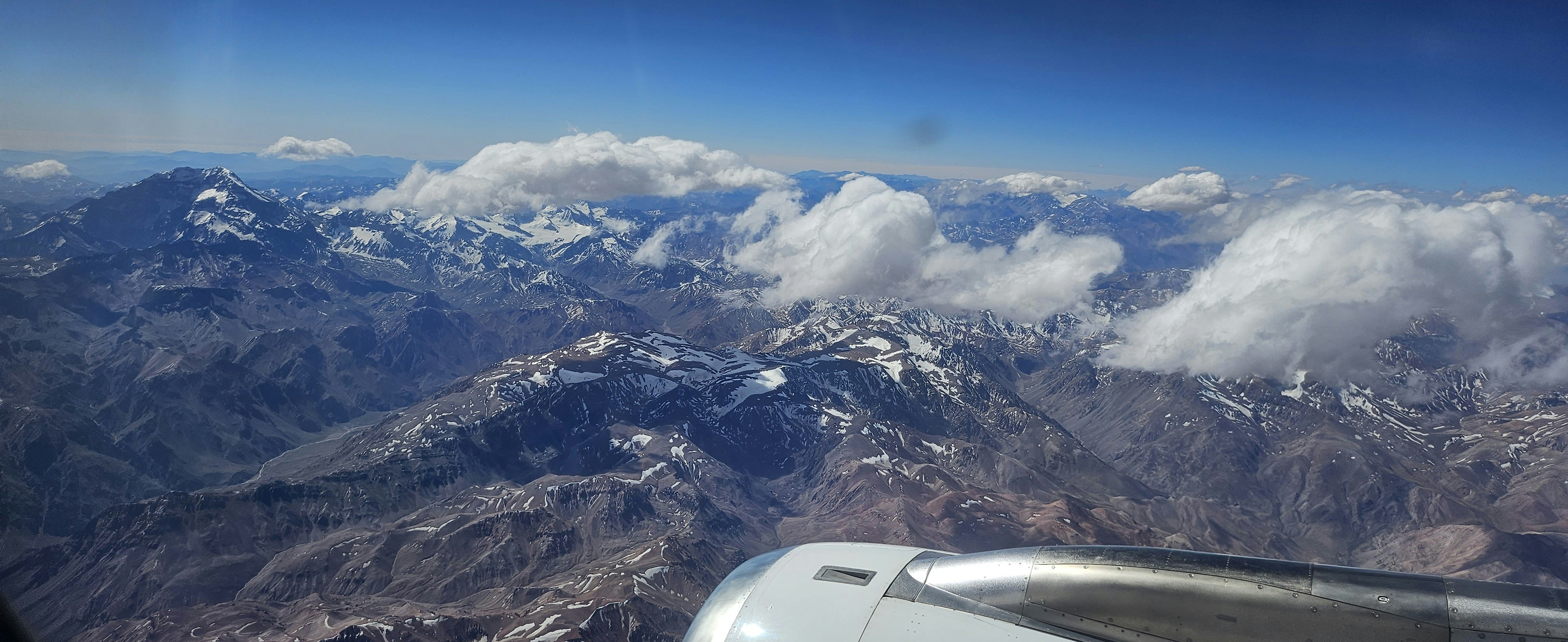 Aerial view of snow-capped mountains with scattered clouds under a clear blue sky.