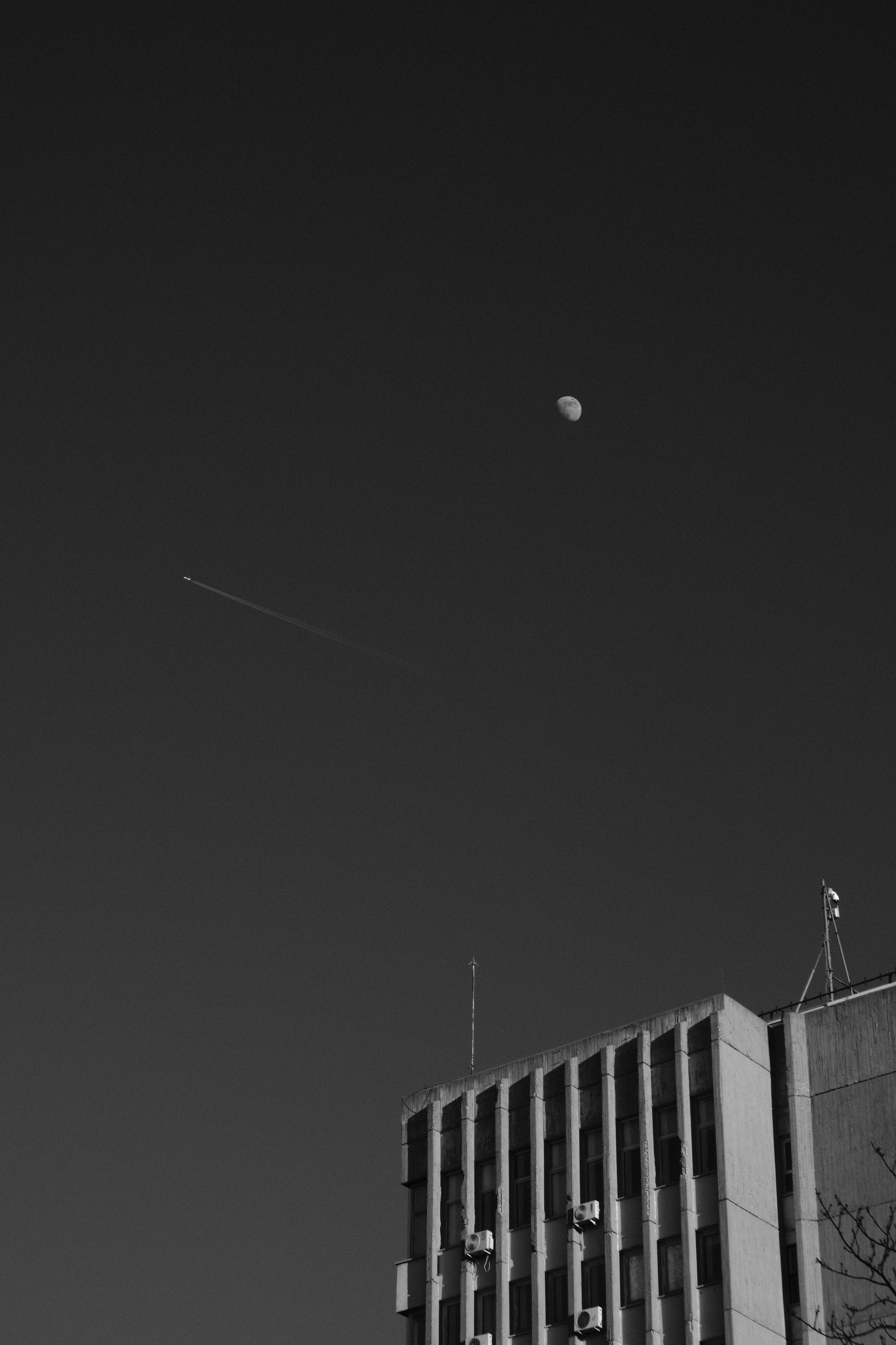 A monochrome scene featuring a tall building with the moon and a contrail above in a clear sky.
