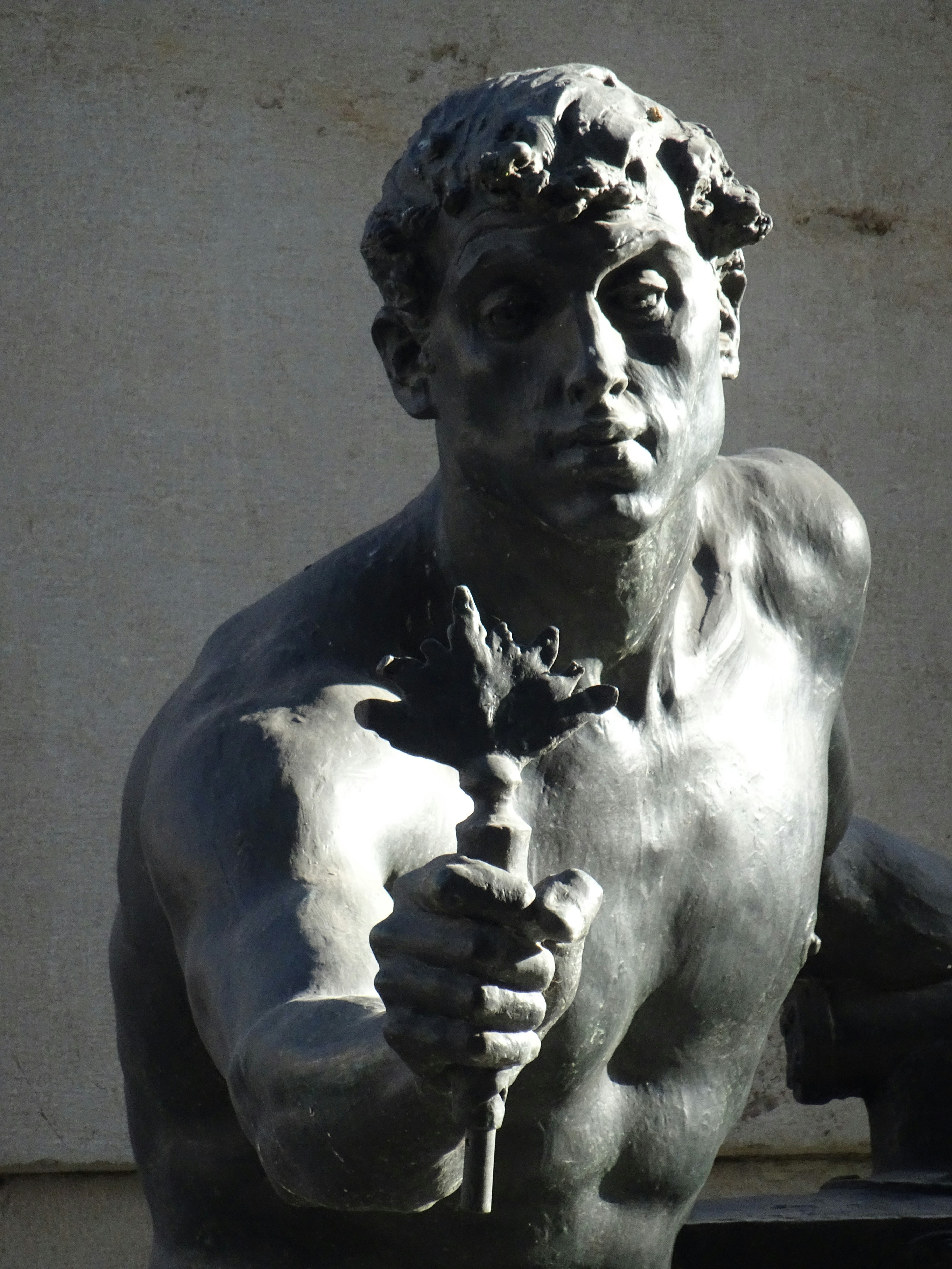 Stone statue bust of a man, his arm extended toward the viewer as he holds a flower-topped staff. Dramatic side lighting emphasizes the sculpted musculature and thoughtful expression.