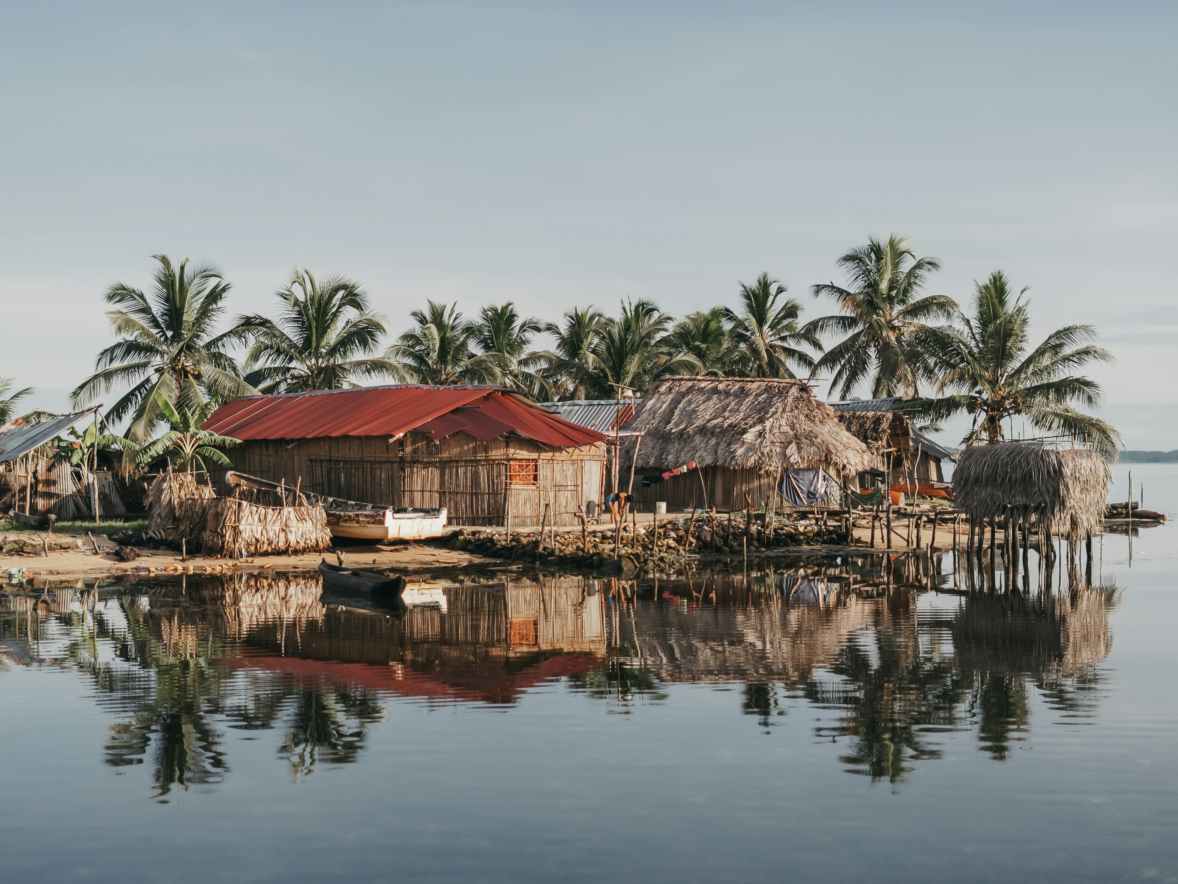 Tropical village by the water with palm trees and thatched-roof homes reflected in calm waters.