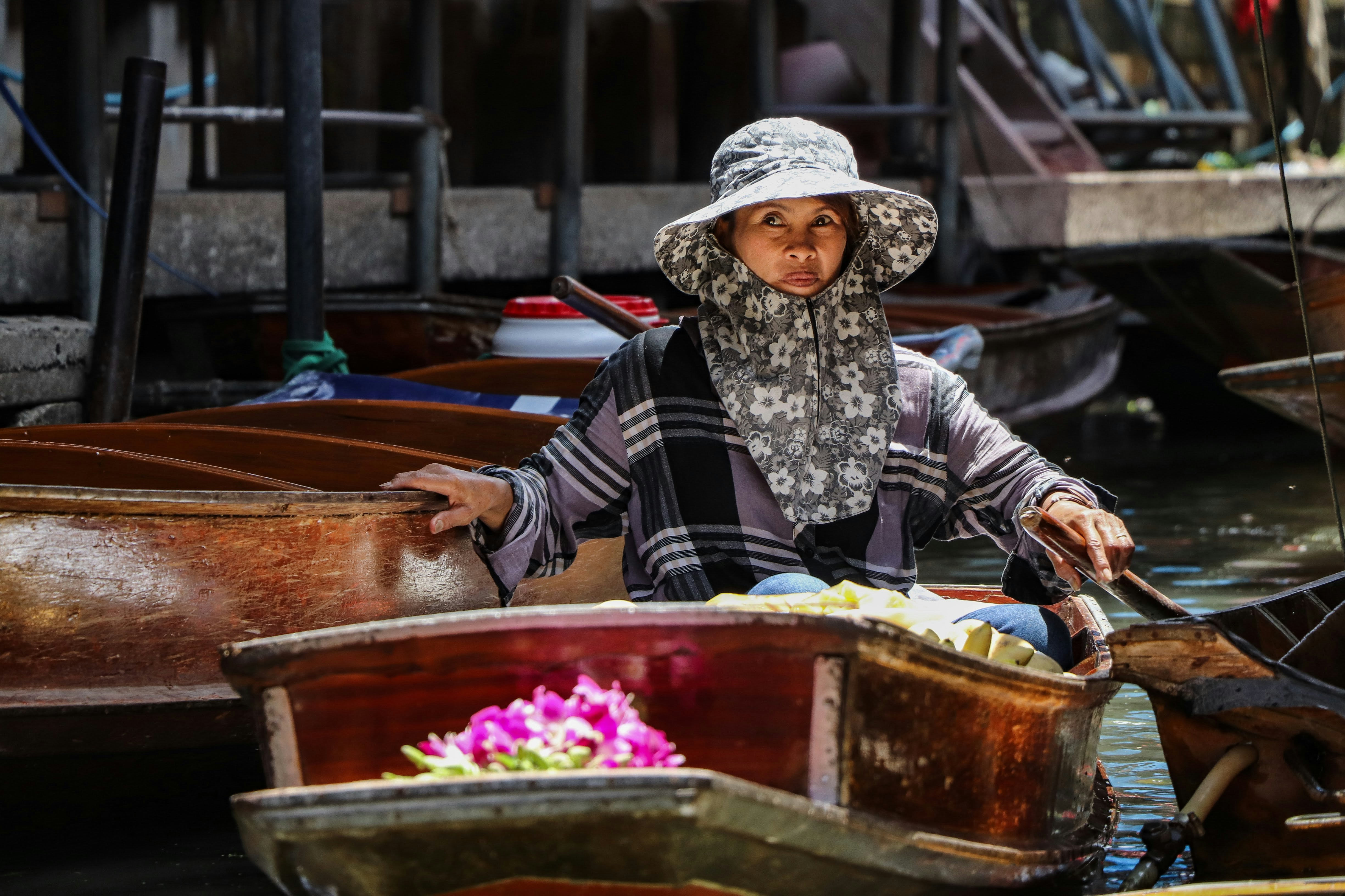 Person in a patterned hat navigates a wooden boat filled with pink flowers at a bustling floating market.
