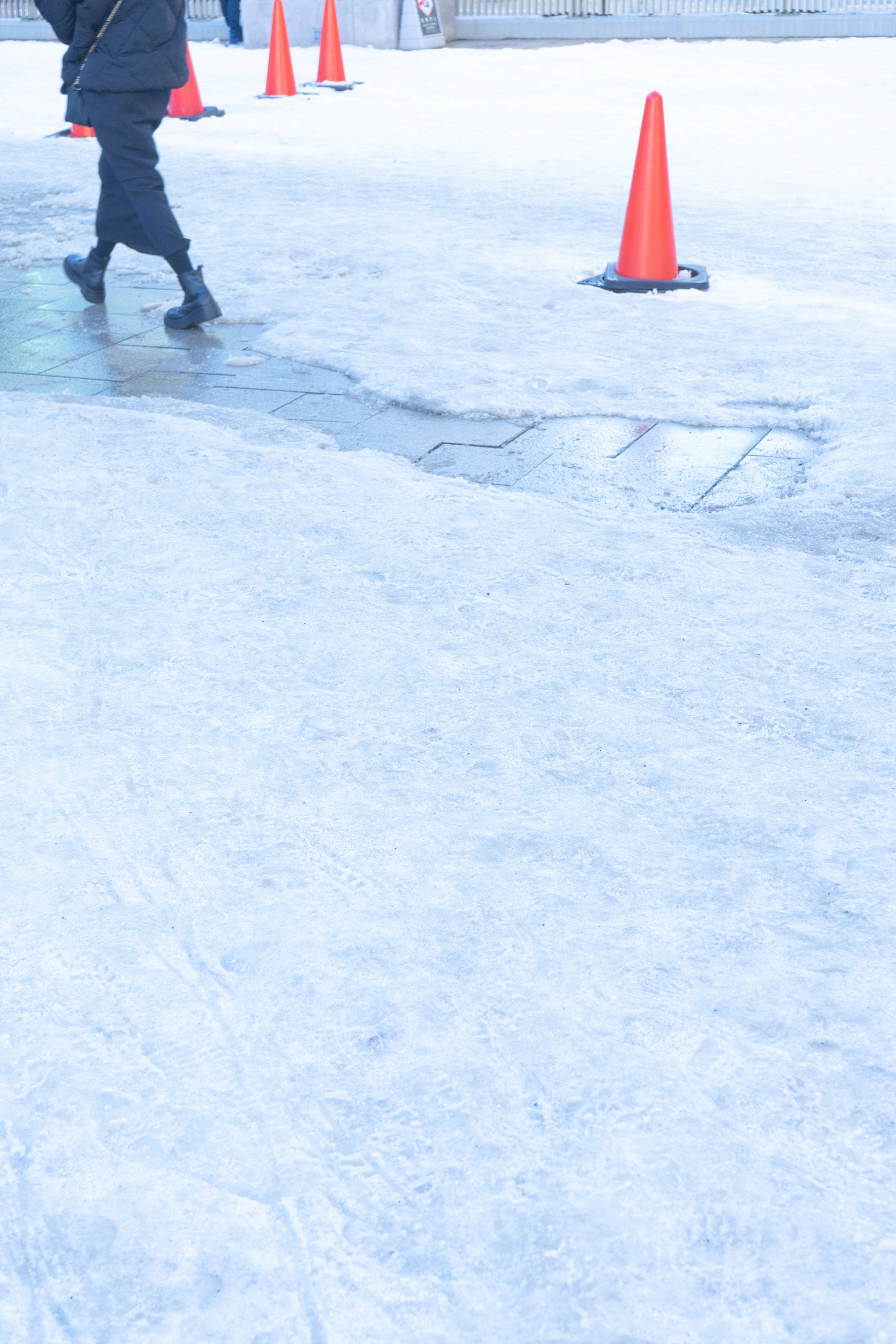 A person in dark clothing walks across a snowy surface, with bright orange cones marking the path. The scene captures the essence of winter navigation.